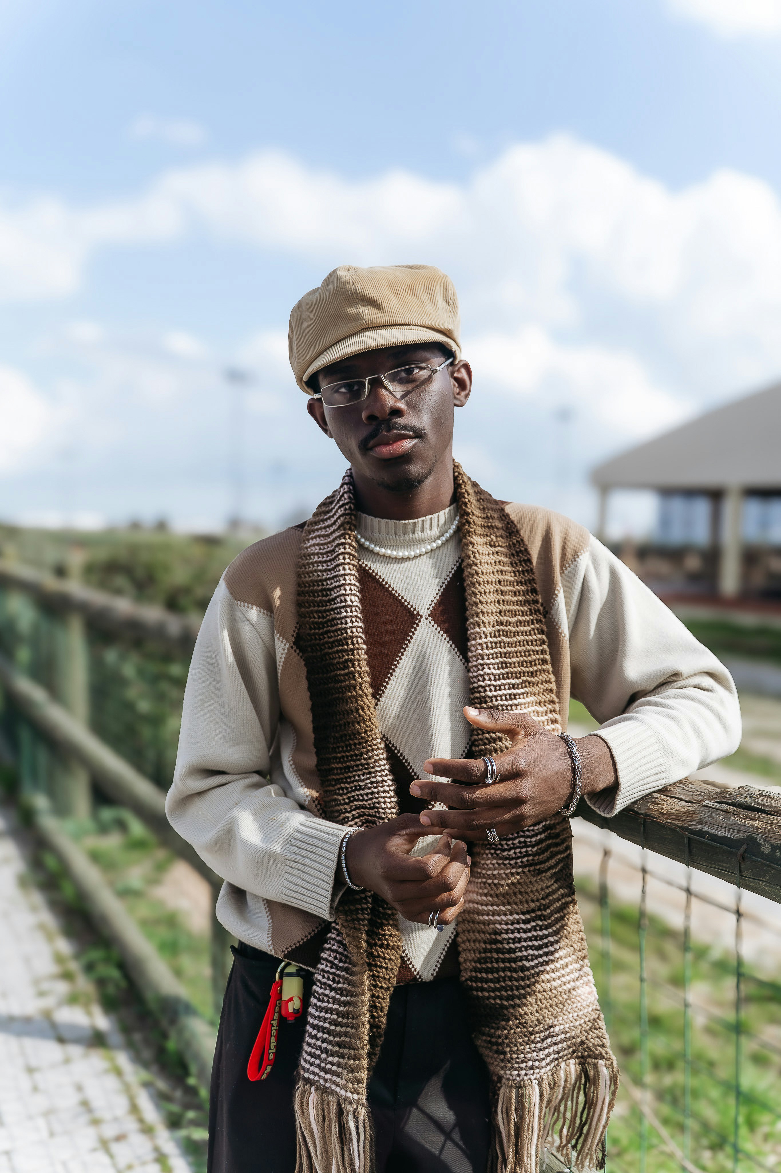 A close-up outdoor portrait featuring a person in vintage-inspired fashion, including a neutral-toned sweater, textured scarf, and cap. Captured with natural light and a soft background, the image highlights modern style mixed with classic, earthy colors.