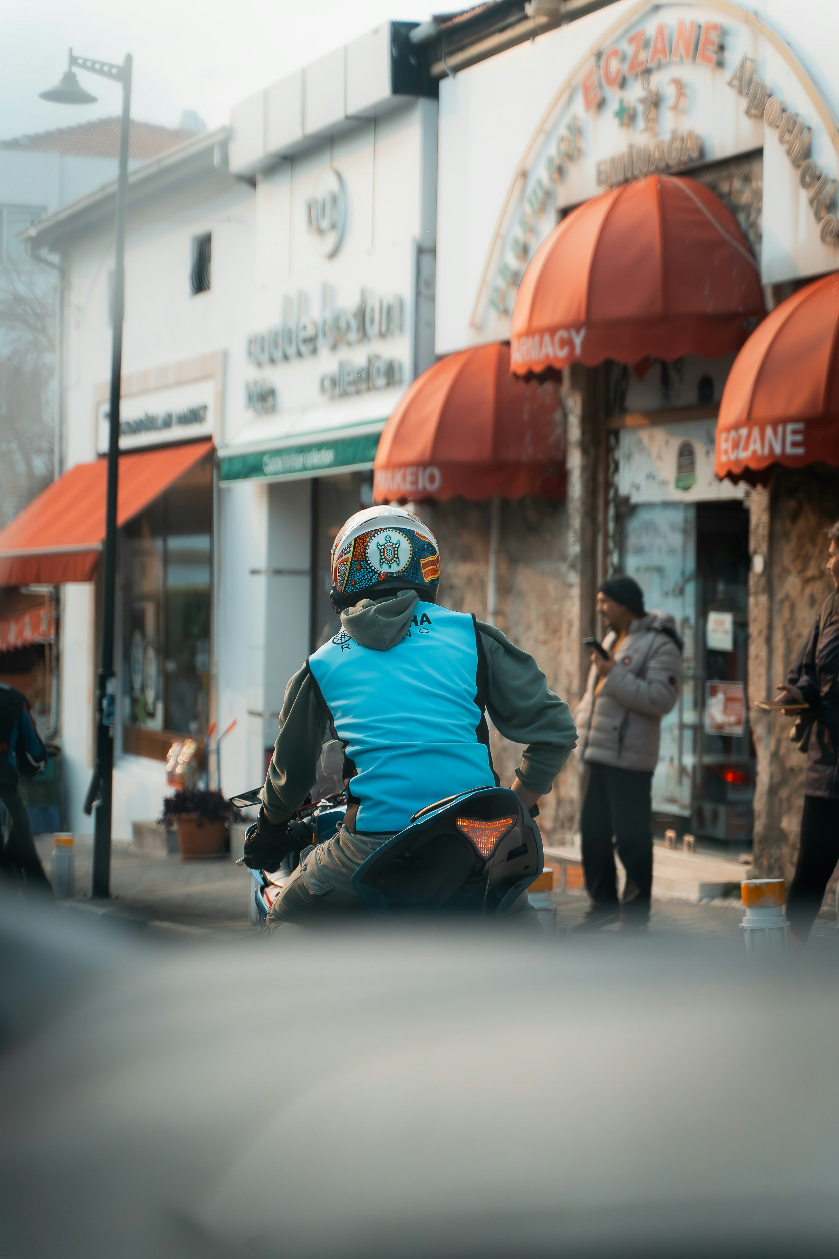 Motorcyclists ride through a narrow urban street lined with buildings, shops, and hanging lights, captured in a cool, cinematic tone. The scene conveys movement, city atmosphere, and a sense of collective journey in a busy neighborhood.