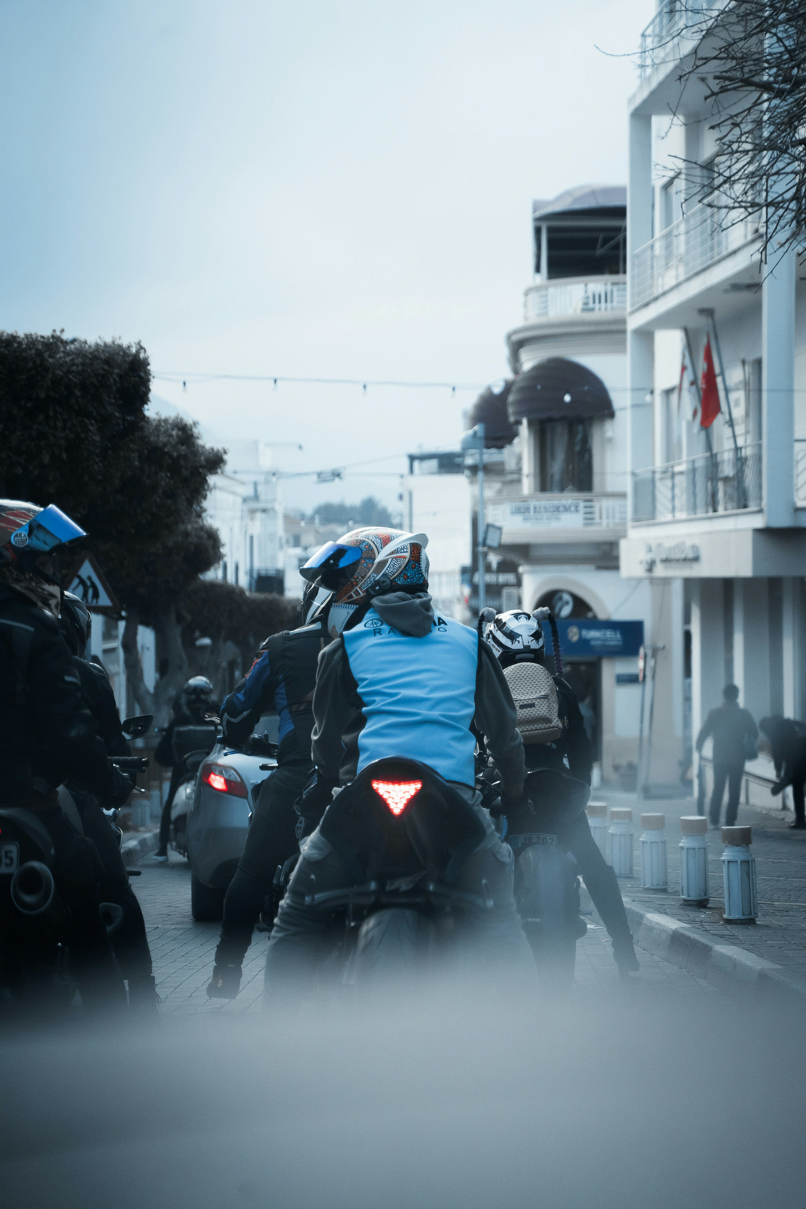 Motorcyclists ride through a narrow urban street lined with buildings, shops, and hanging lights, captured in a cool, cinematic tone. The scene conveys movement, city atmosphere, and a sense of collective journey in a busy neighborhood.