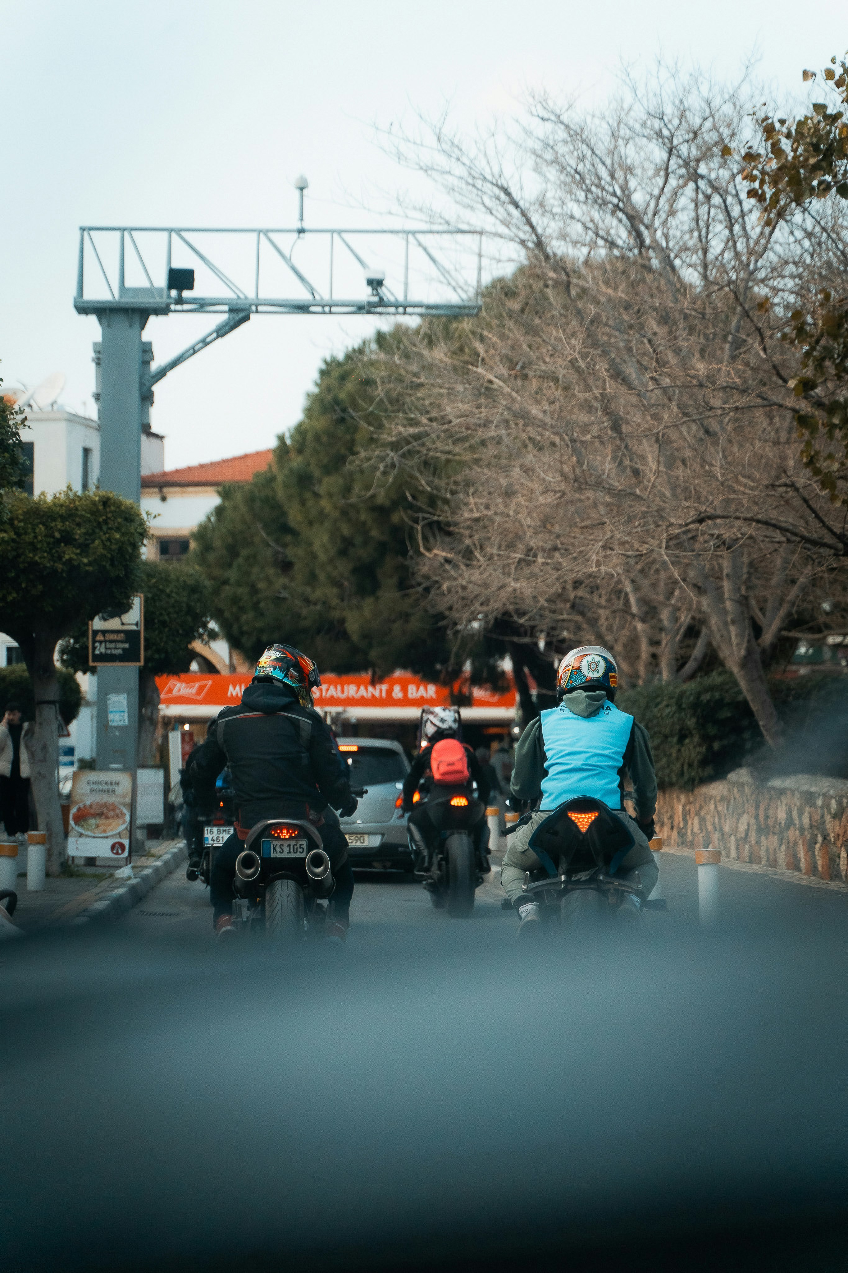 Motorcyclists ride through a narrow urban street lined with buildings, shops, and hanging lights, captured in a cool, cinematic tone. The scene conveys movement, city atmosphere, and a sense of collective journey in a busy neighborhood.