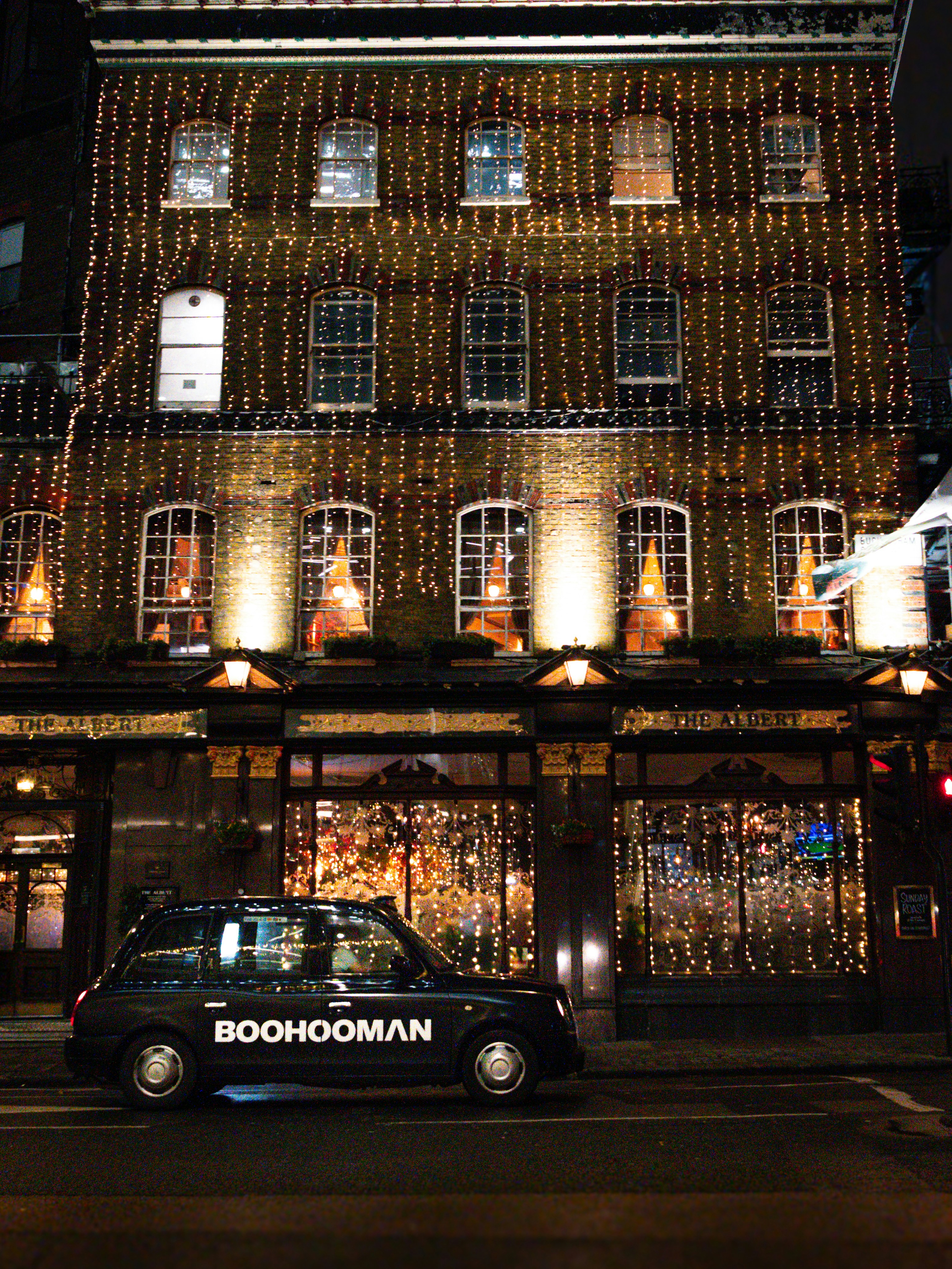 Nighttime Brilliance: The BoohooMan Taxi in Front of a Festive Brick Facade