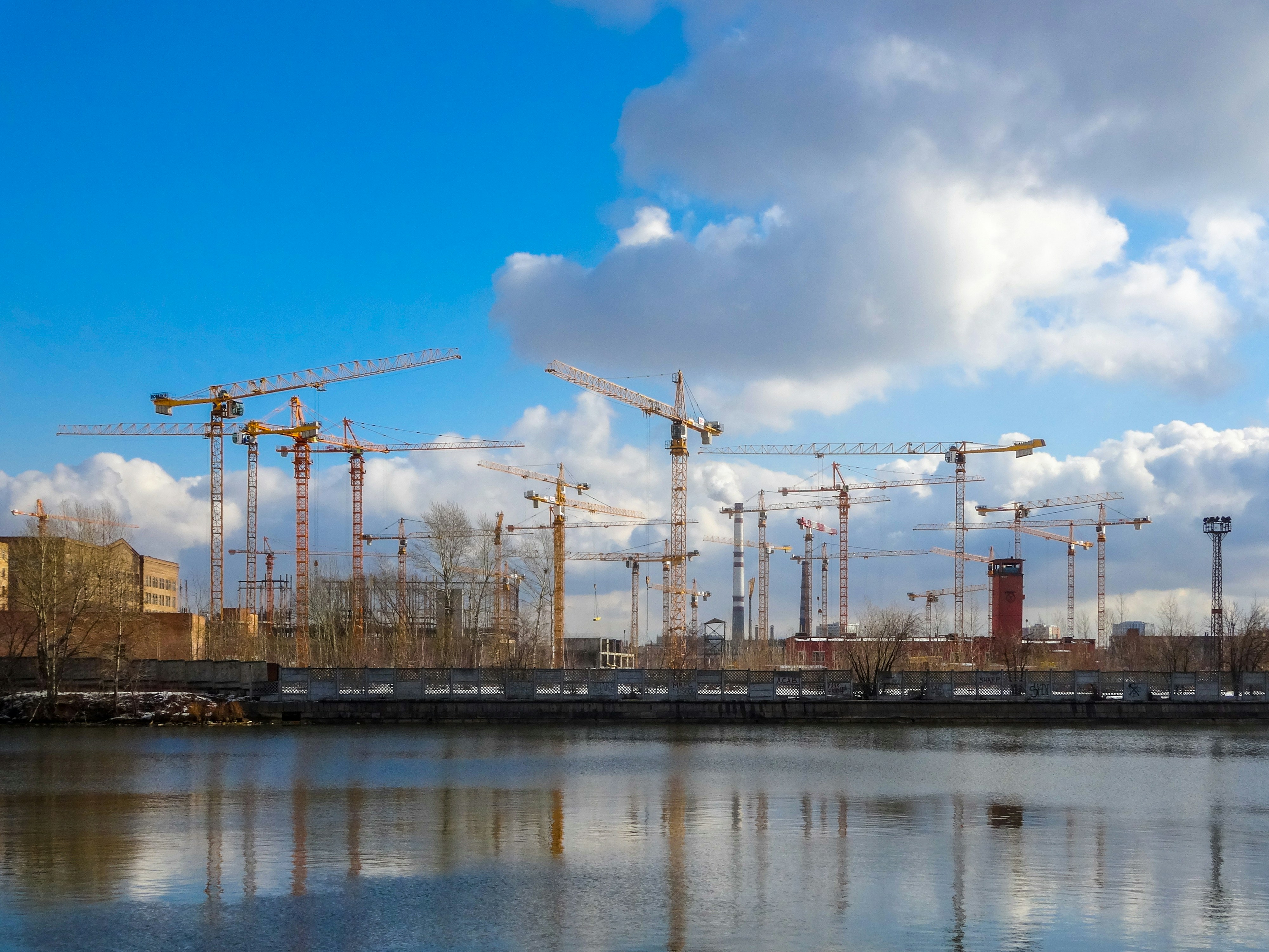 Construction cranes by the water under a cloudy sky.