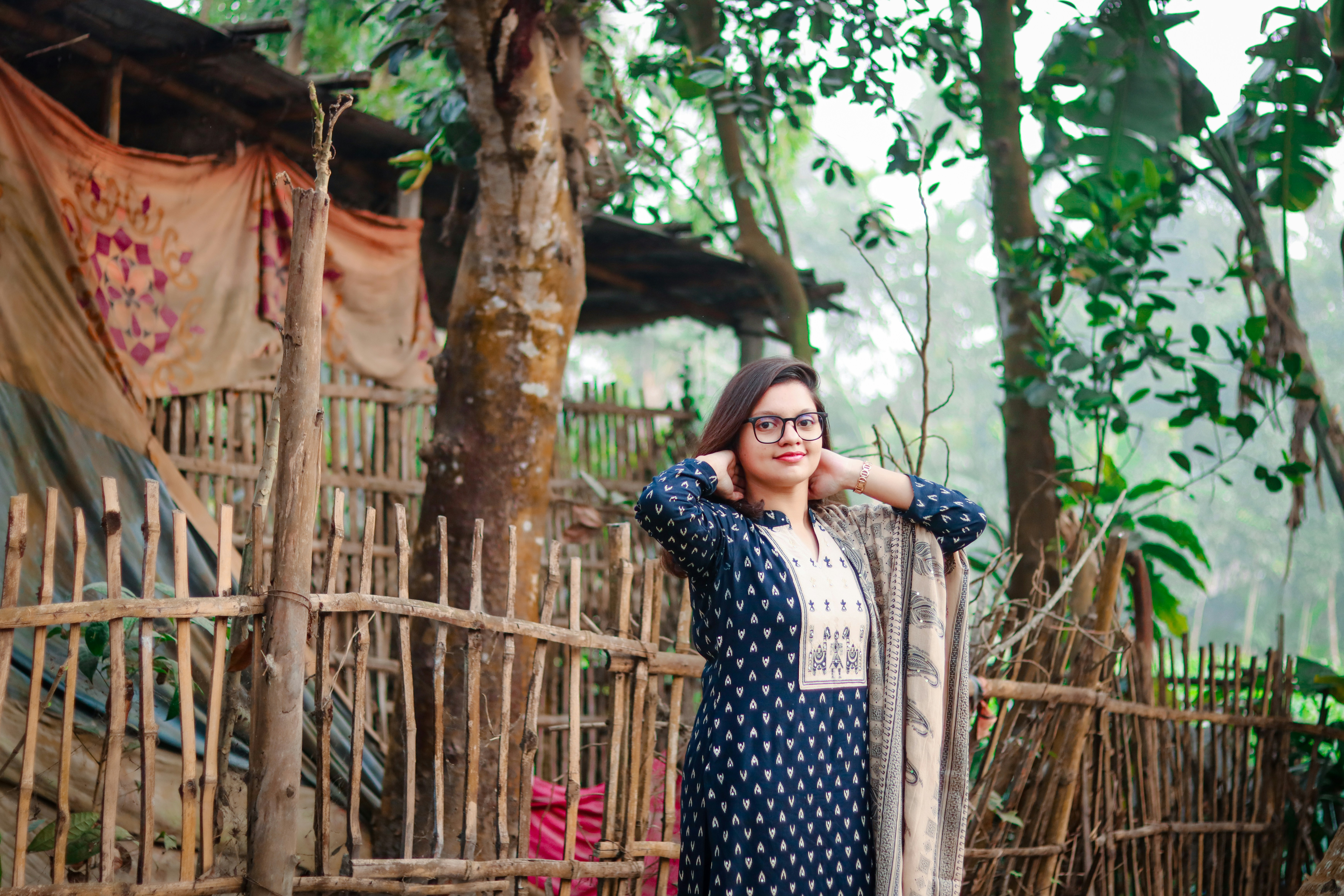 Woman in patterned dress stands before rustic bamboo shelter in a verdant setting