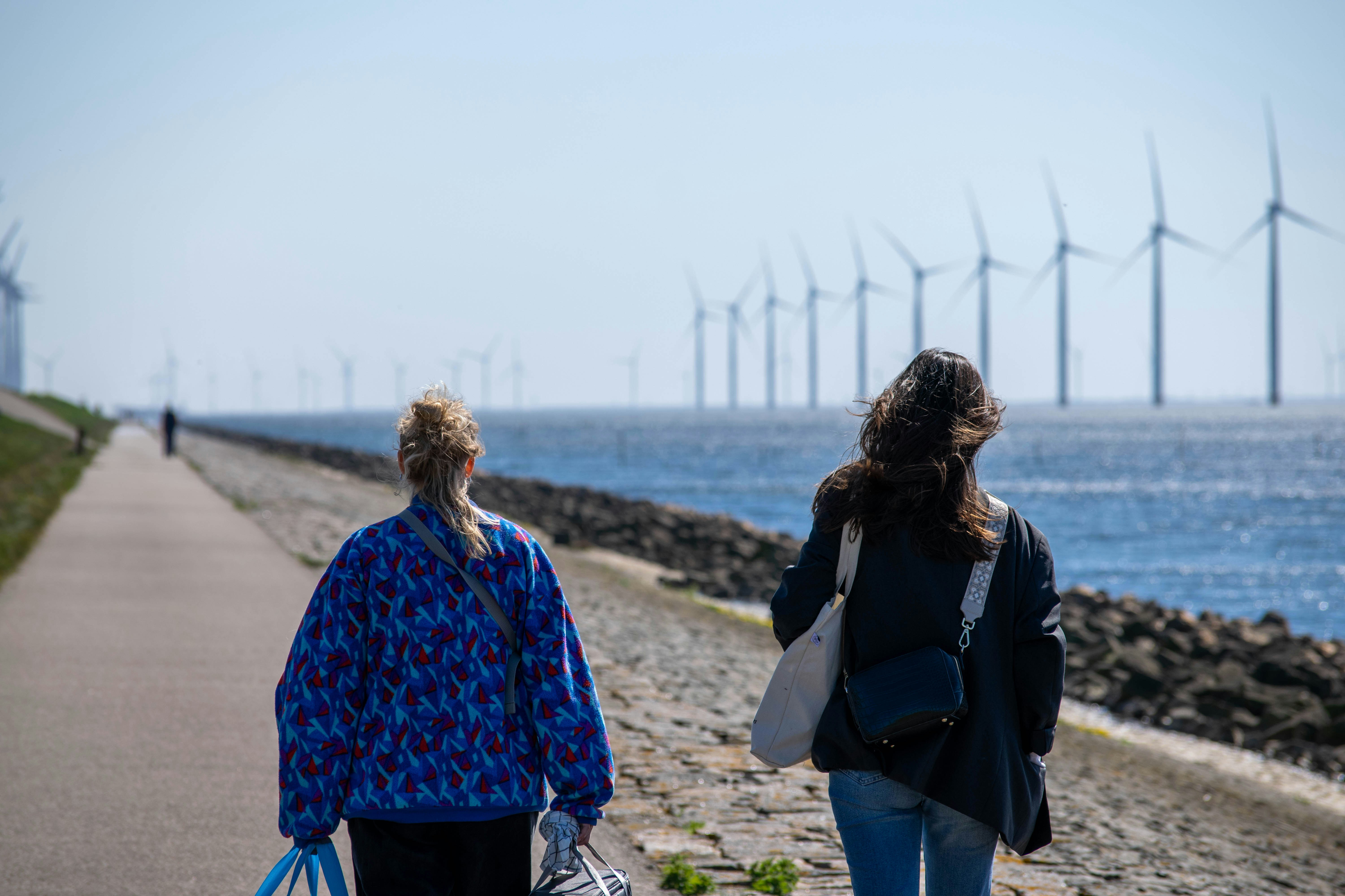 Two pedestrians stride along a windy coastal promenade past a menacing line of offshore wind turbines