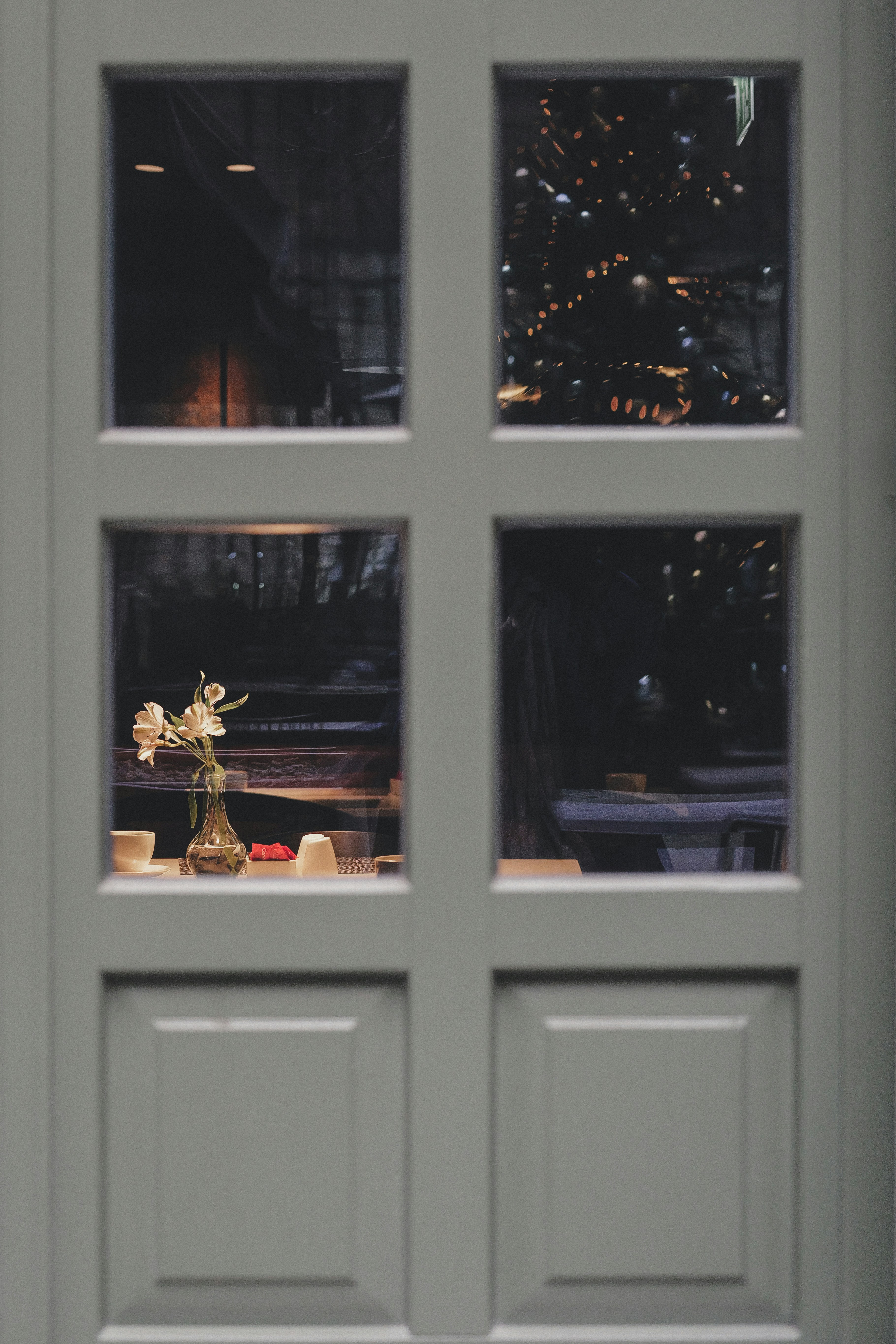 A quiet street-side window, a vase of white lilies glows against a dark interior, framed by muted grey panels and soft evening reflections.
