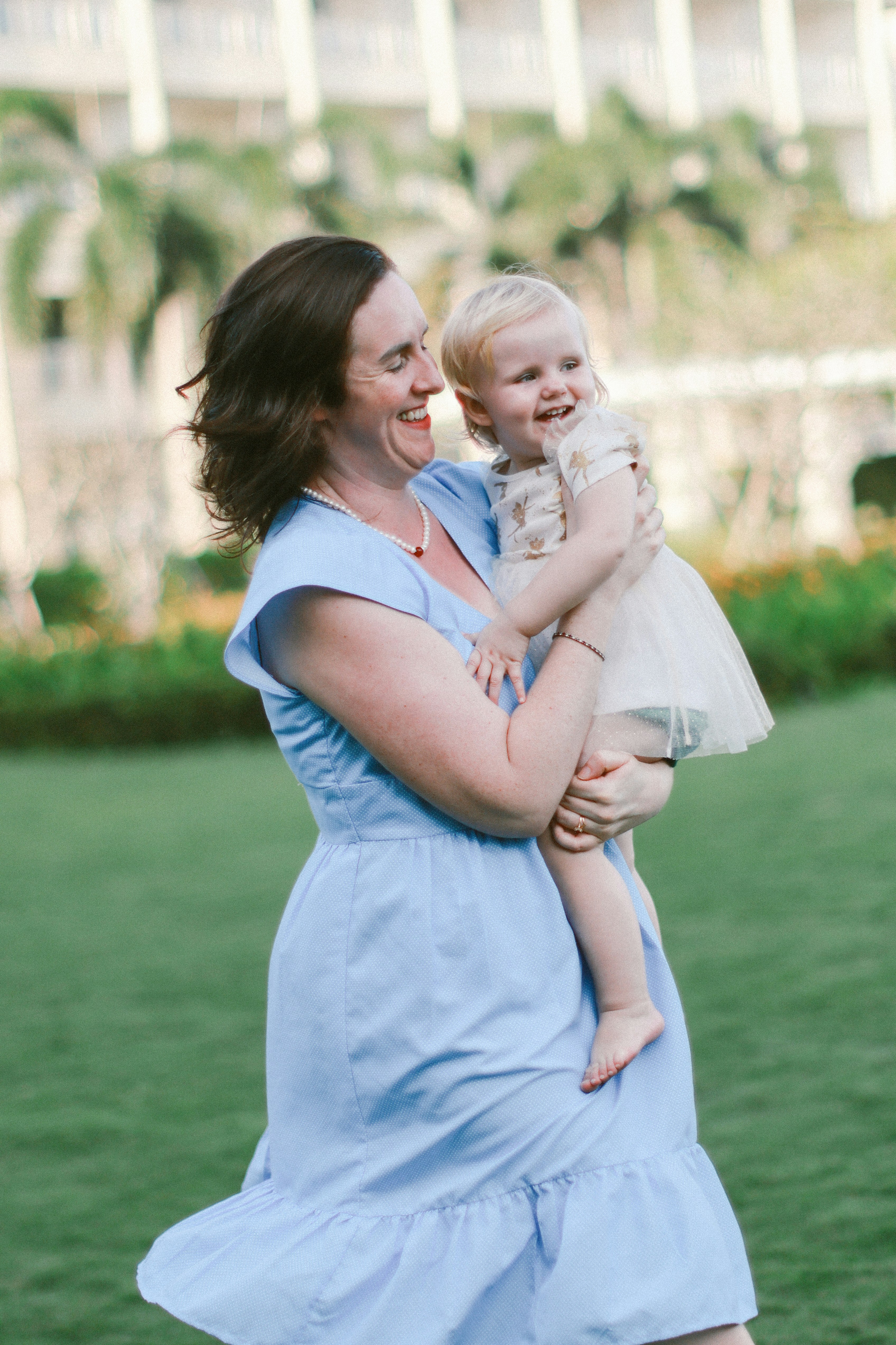 Mother in a sky-blue dress lifts joyful baby in a sunlit garden