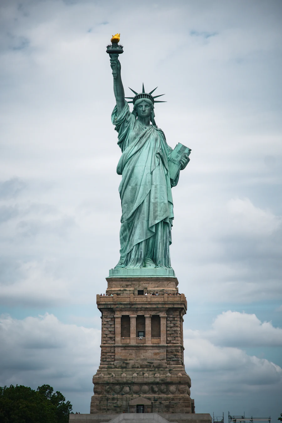Bronze-green Statue of Liberty rises atop a sandstone pedestal against a cloudy sky, framed for a classic cityscape portrait.