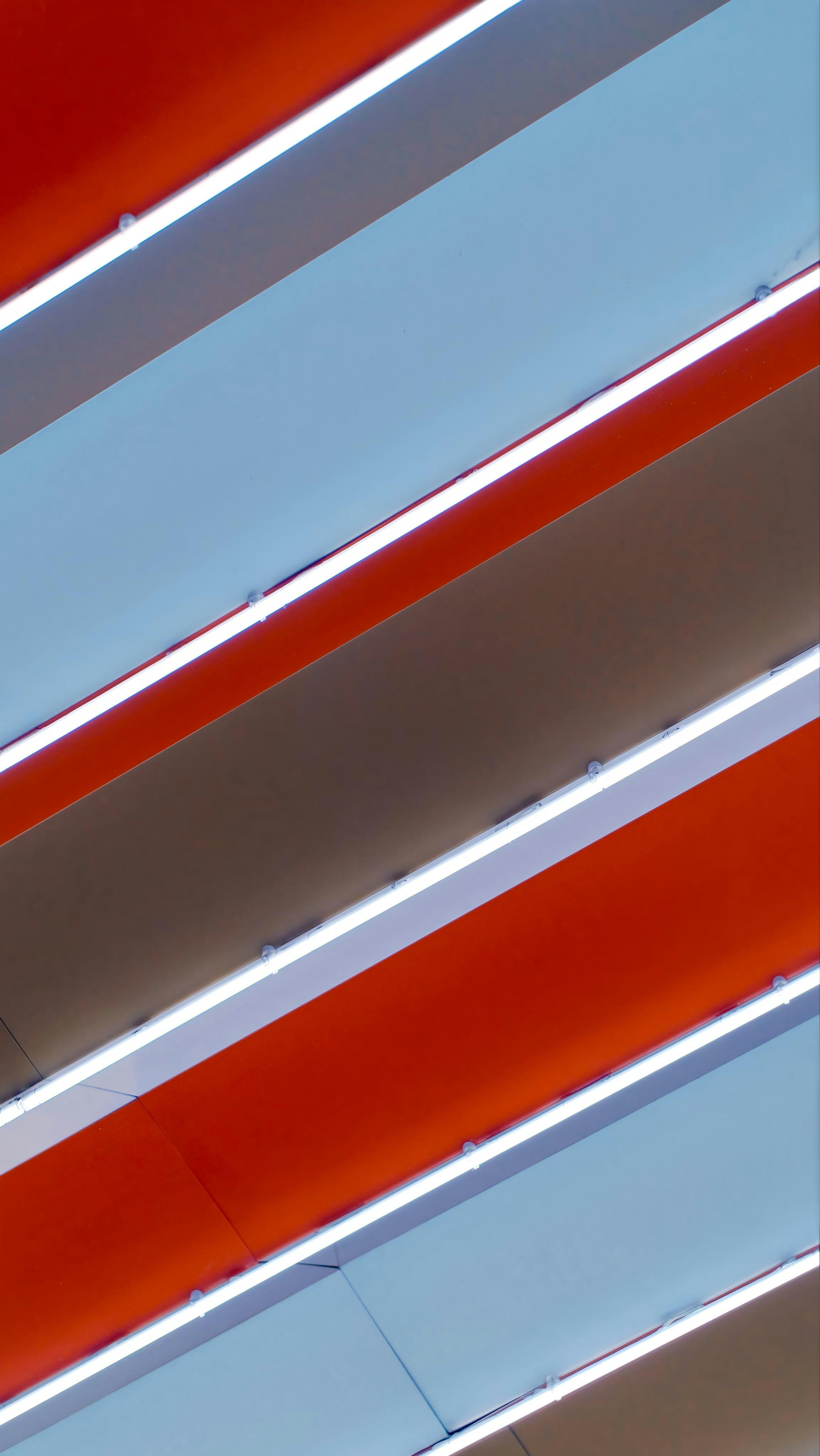 Abstract architectural composition of colorful ceiling panels and fluorescent lights forming strong diagonal lines. A vibrant mix of red, blue, and neutral tones creates a modern geometric pattern with rhythm, contrast, and symmetry. Perfect as a background for design, branding, technology, and creative projects