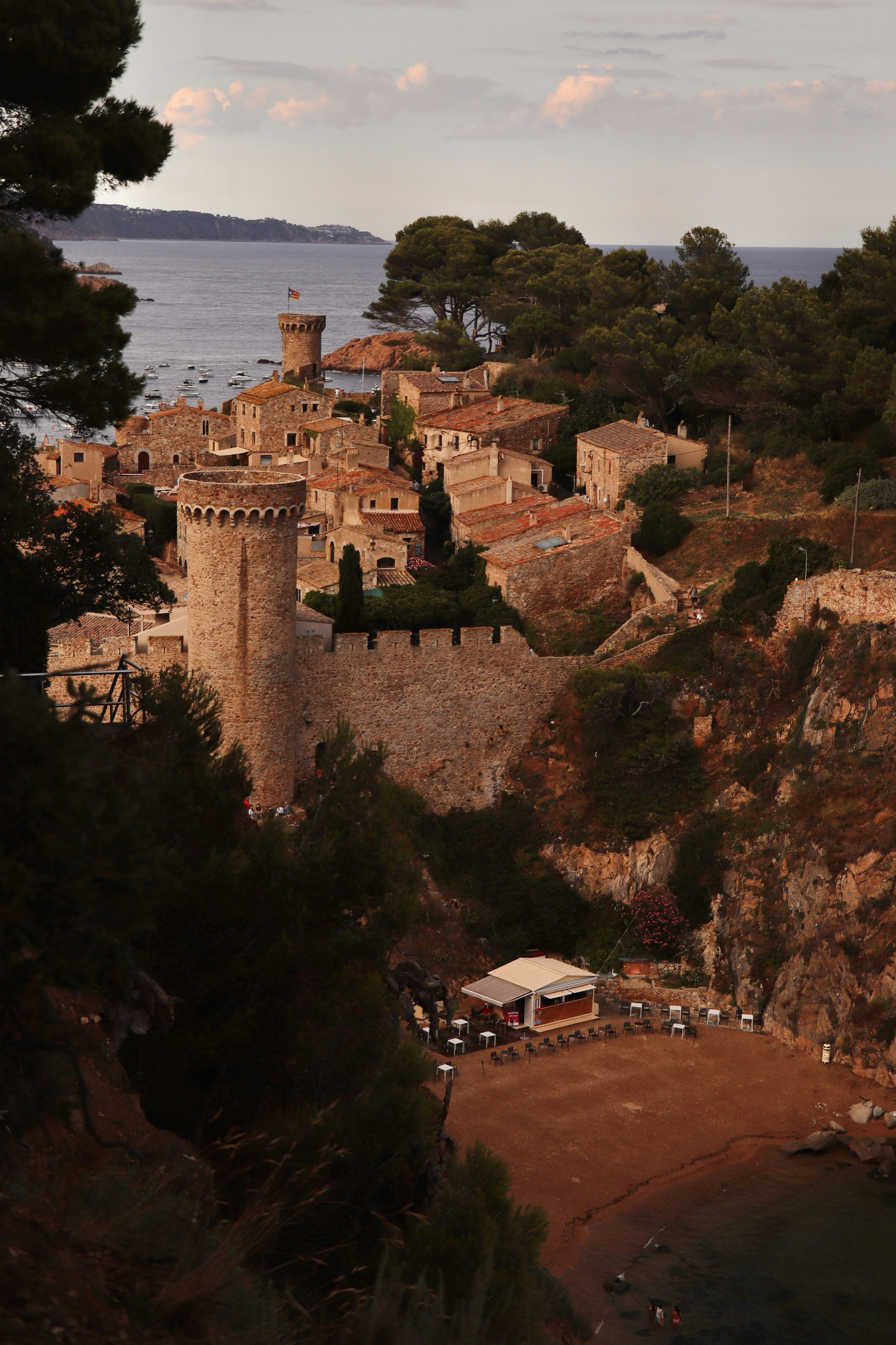 Golden hour over Vila Vella in Tossa de Mar, Catalonia: a round medieval watchtower and crenellated walls rise above a secluded sandy cove with a small beach bar, terracotta rooftops and pine-covered cliffs meeting the calm Mediterranean. A classic Costa Brava scene that blends history, nature and seaside life.