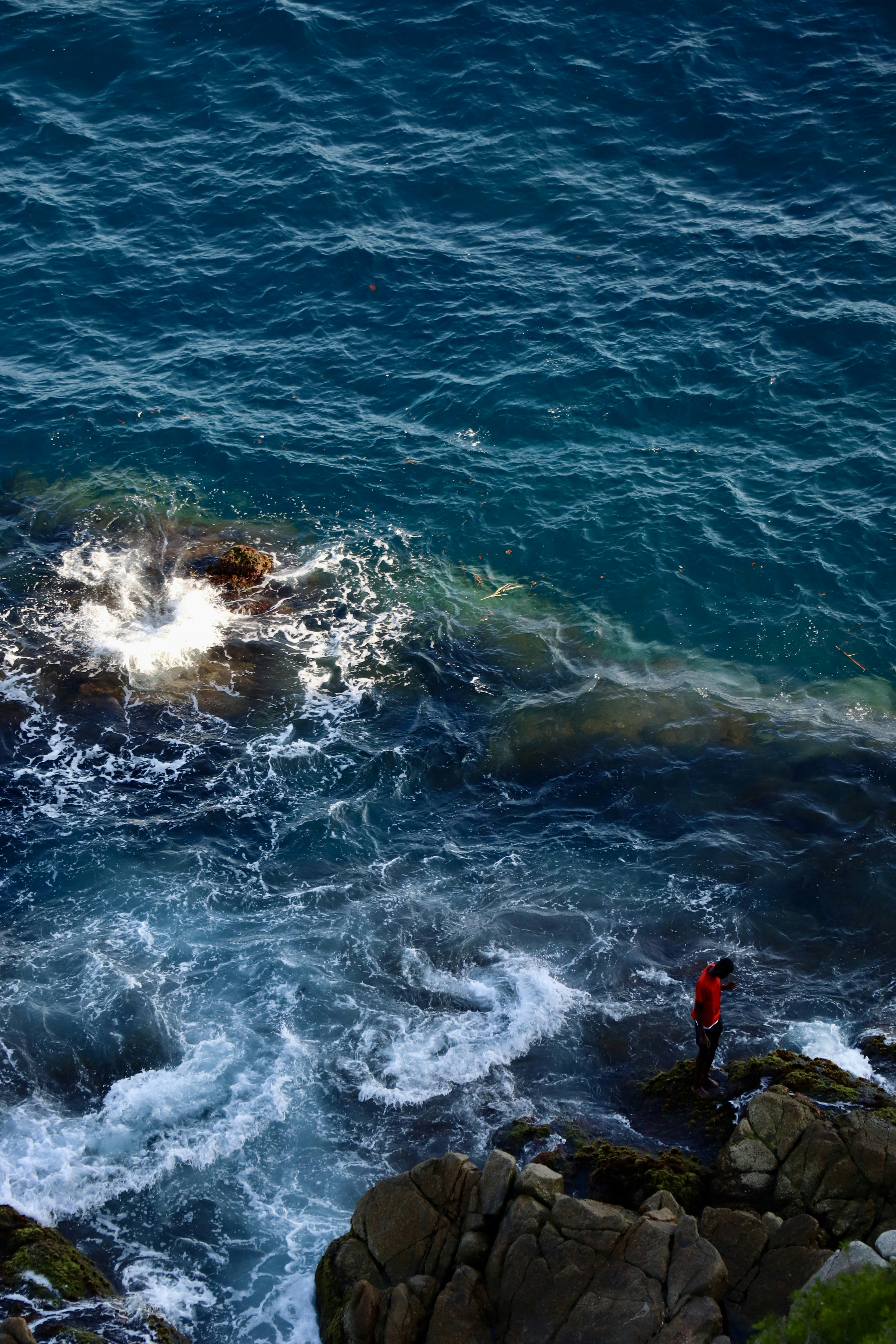 A lone figure in a red jacket stands on slick coastal rocks as teal waves surge and fizz around them. The frame is mostly sea, textures of ripples, foam and spray, so the person feels small, adding a quiet sense of scale, solitude and contemplation.