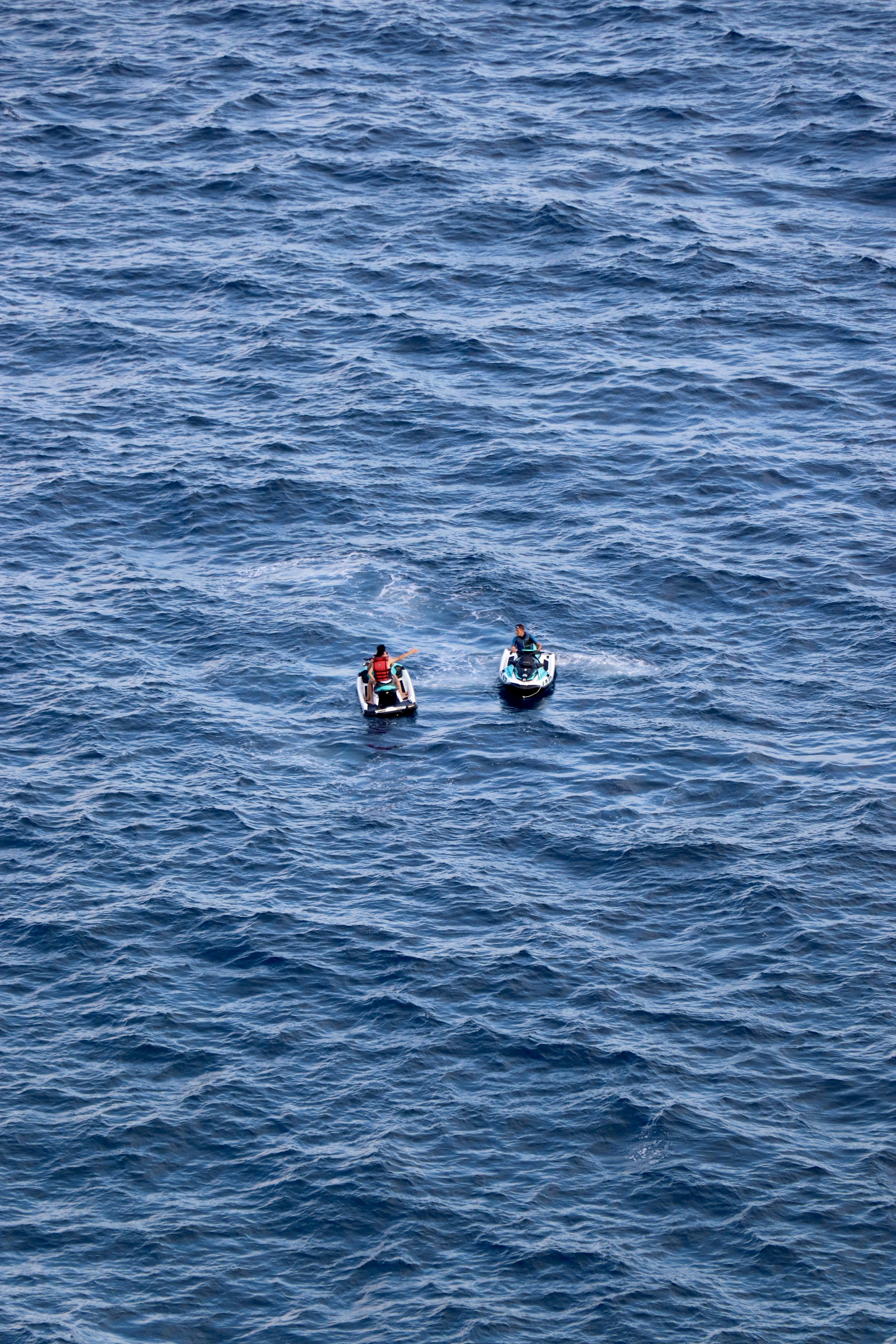 Two riders on personal watercraft cross the open sea, framed by a vast expanse of textured blue waves. The high-angle view and generous negative space highlight motion, scale, and a minimalist summer vibe.