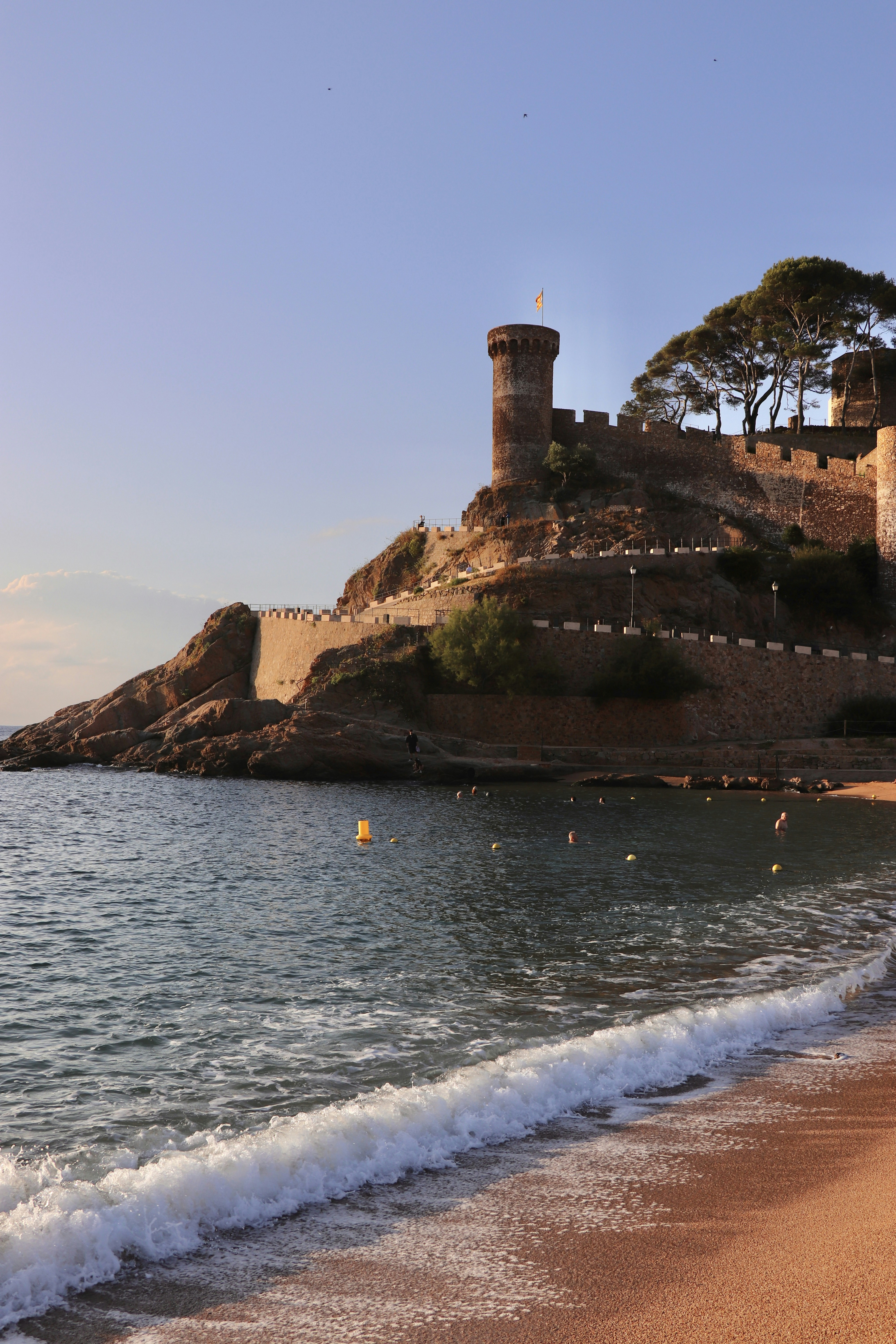 Golden-hour view of the medieval castle tower and stepped ramparts of Tossa de Mar on the Costa Brava, with small waves washing over the sandy beach and a calm Mediterranean cove below.