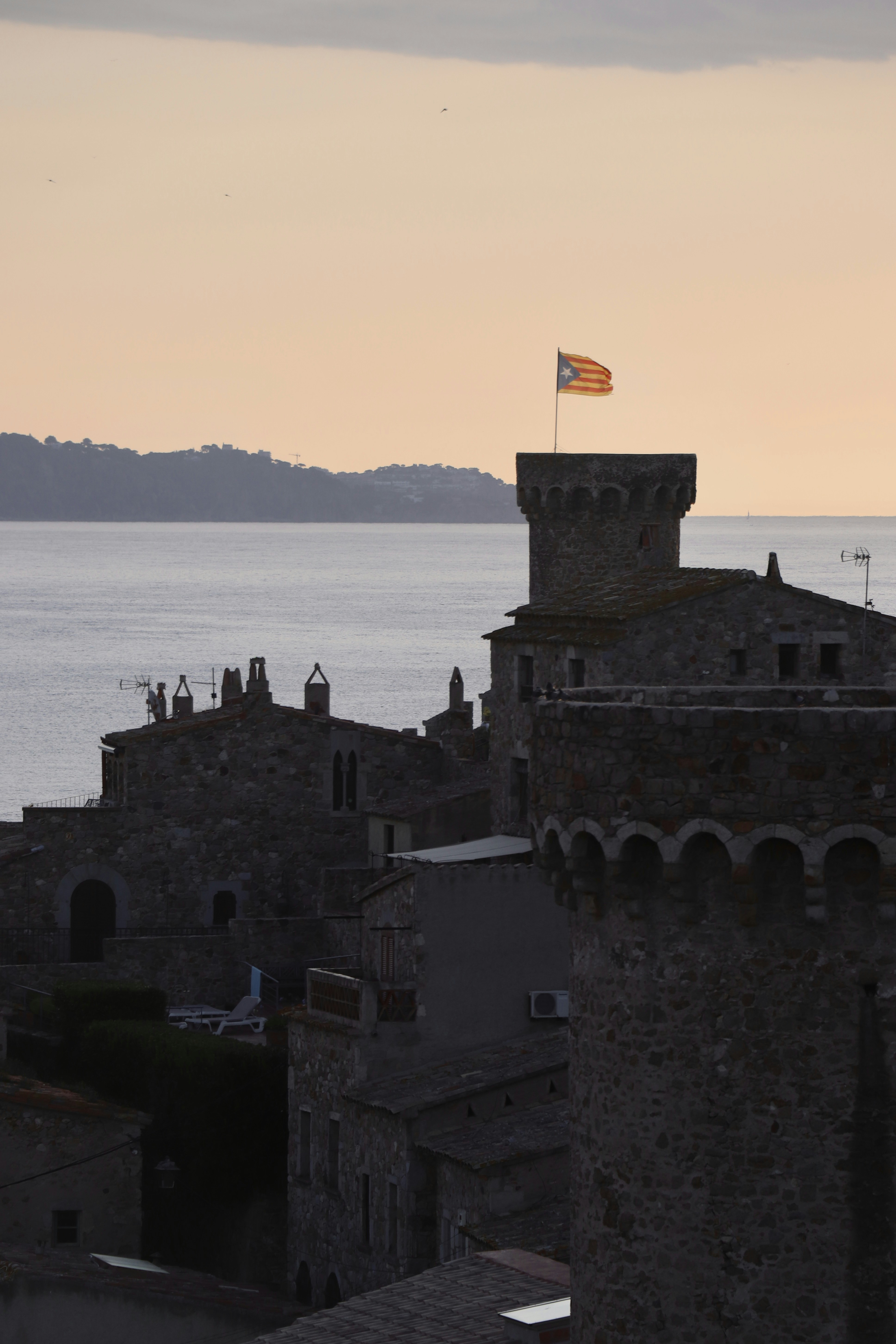 Silhouette of the medieval stone towers of Tossa de Mar at sunset, a Catalan flag fluttering above the ramparts as warm dusk light fades over the calm Mediterranean Sea.