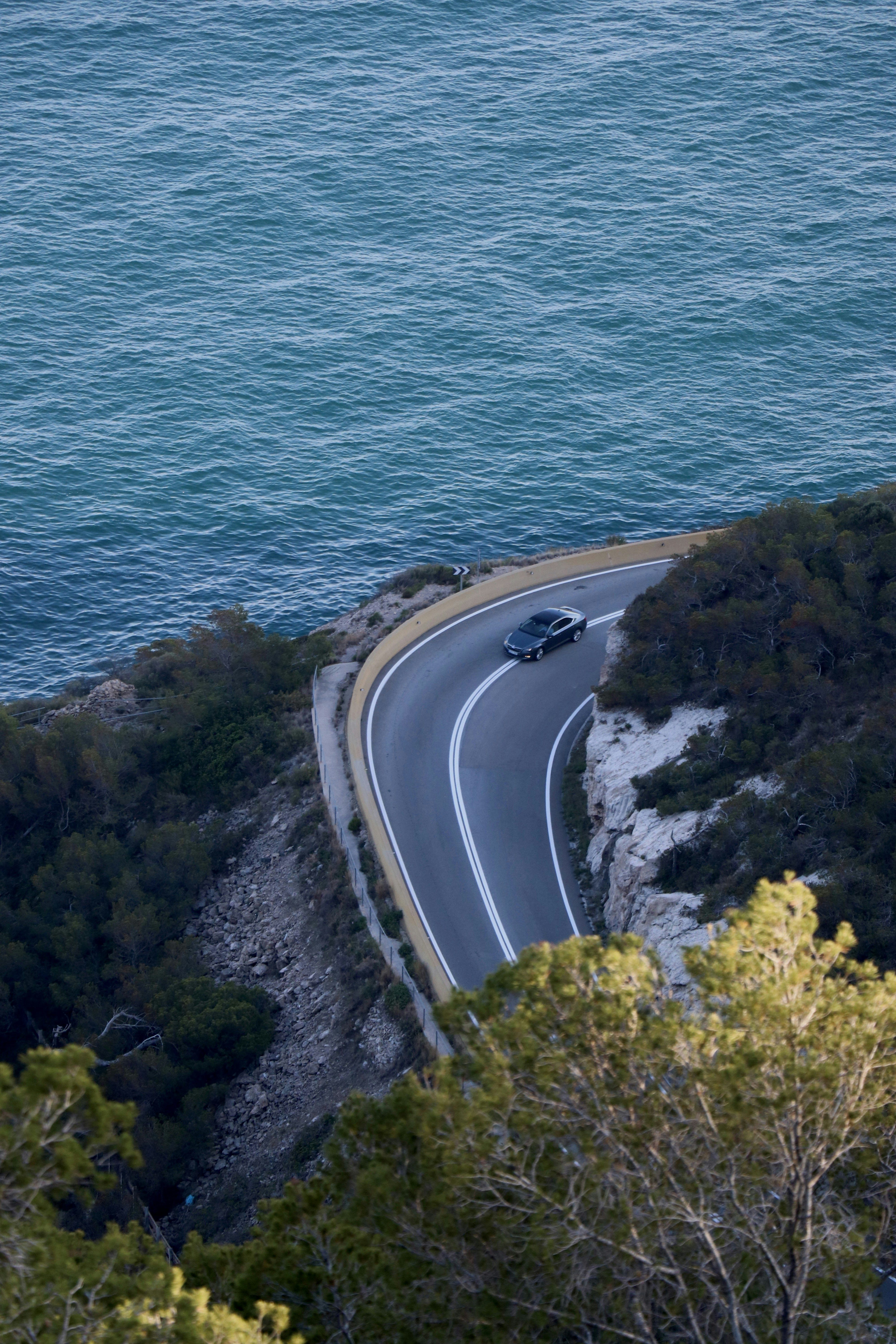 A single car navigates a tight switchback on a scenic coastal road, framed by cliffs and turquoise sea, airy, and made for road-trip vibes.