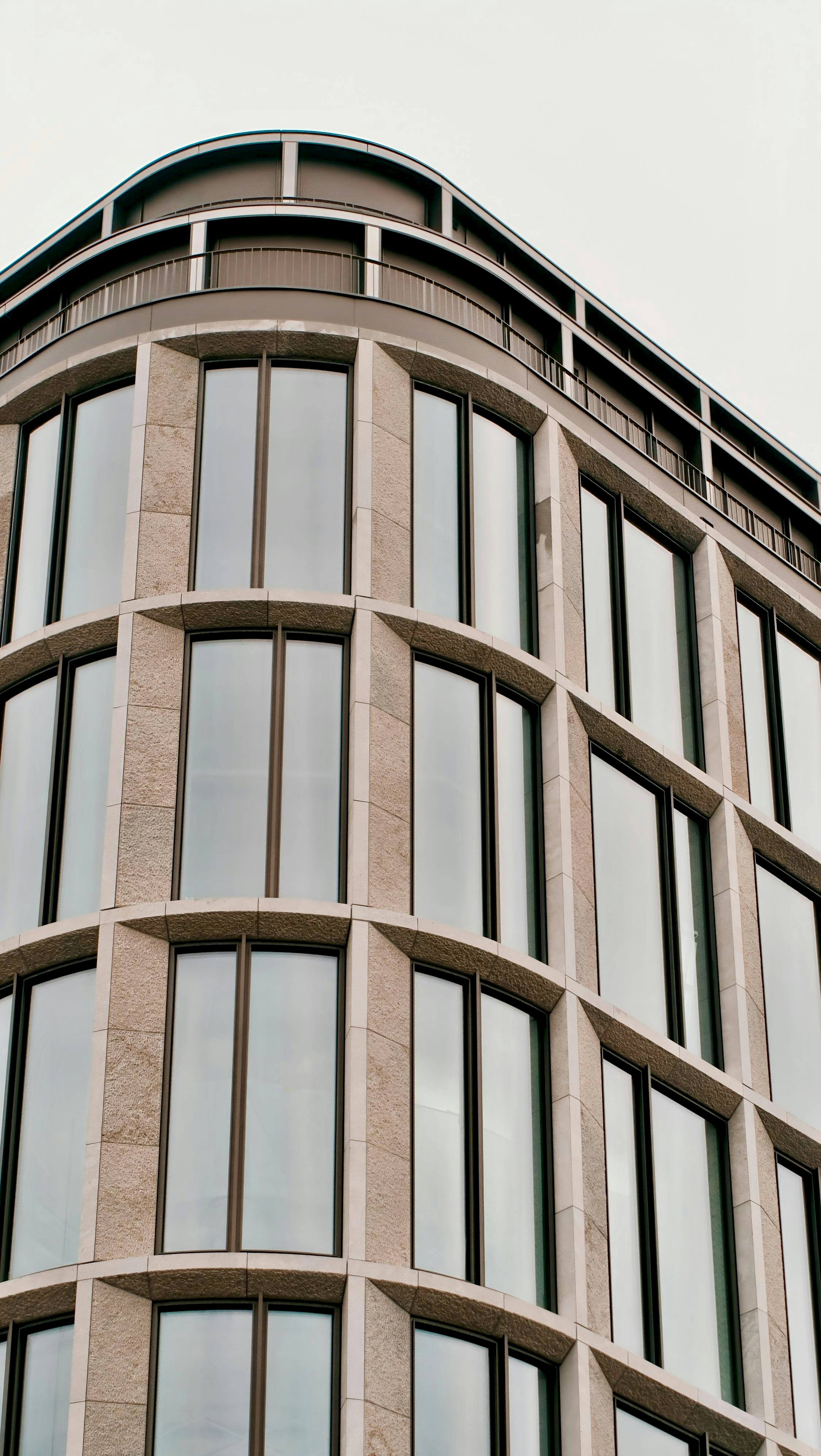 The curving, textured facade of a modern corporate building, featuring a repeating pattern of large, recessed glass windows and beige stone framework under a soft, overcast sky. The rounded corner emphasizes the architectural symmetry and contemporary design.