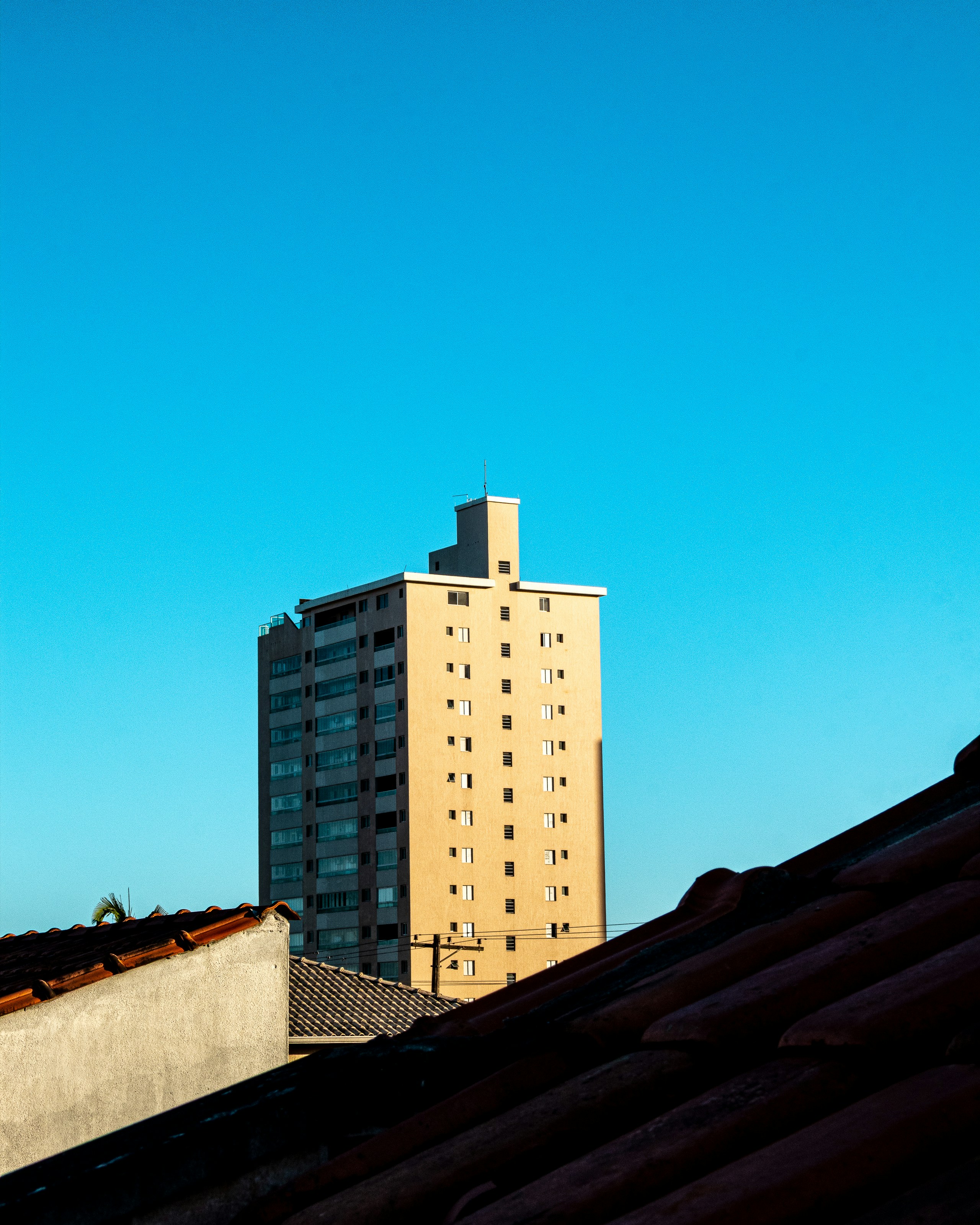 A tall, tan apartment building stands against a clear blue sky, with the foreground showing the tops of tiled roofs and a white wall.