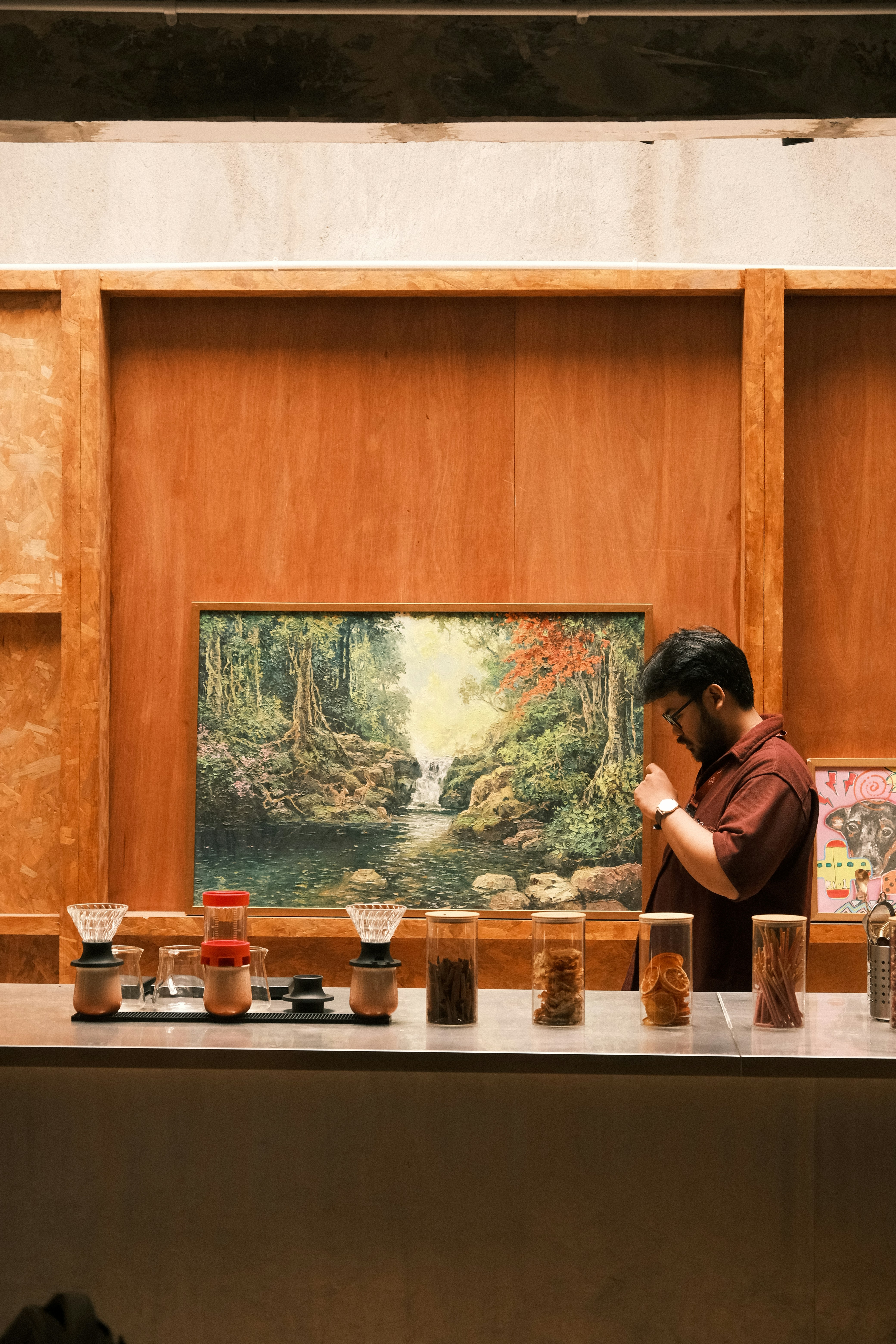 A cozy coffee shop in Jakarta featuring a serene forest landscape painting. The relaxed ambiance is complemented by a barista preparing coffee, creating a perfect environment for unwinding and enjoying art and coffee.