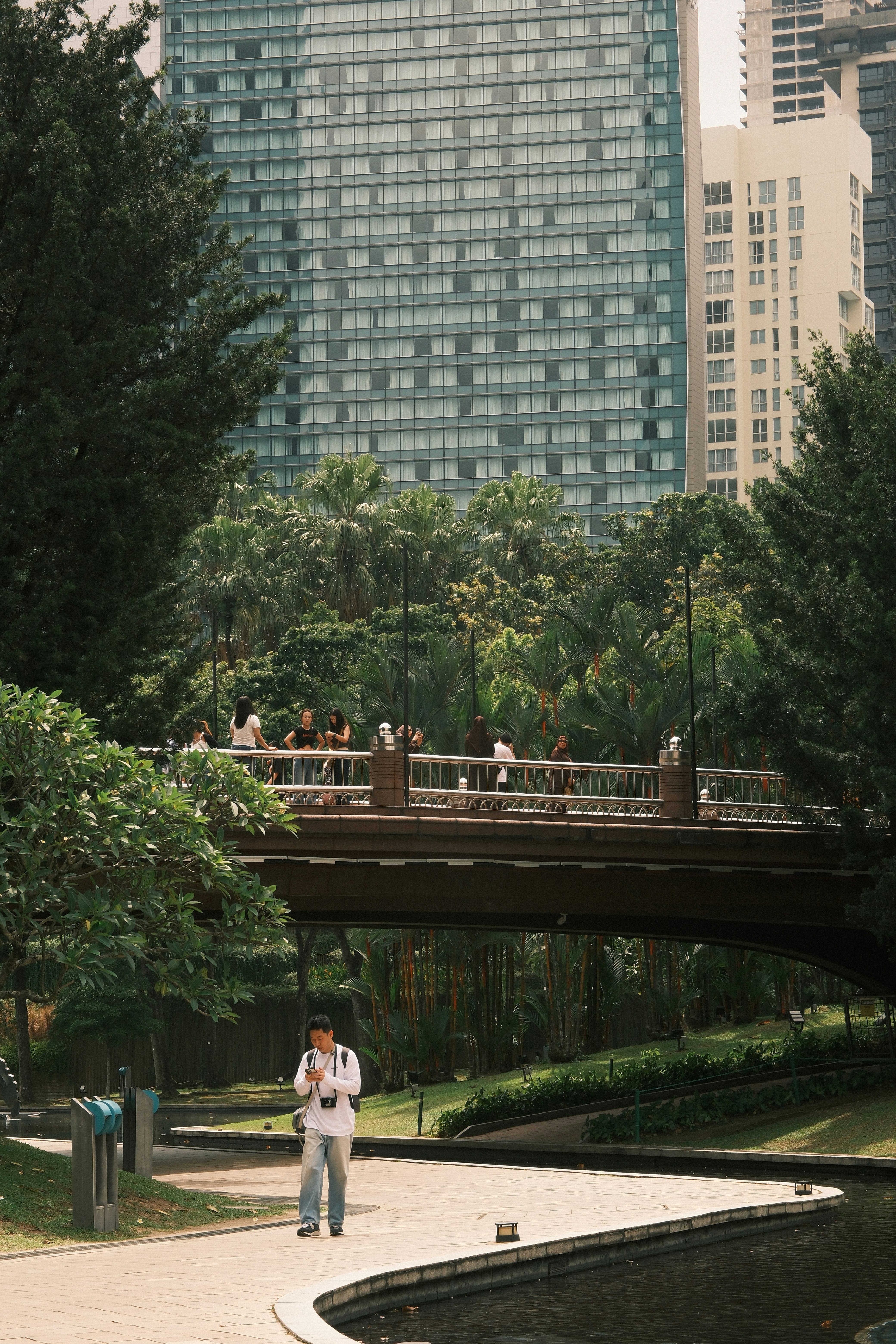 A serene scene at KLCC Park in Kuala Lumpur, with people crossing a bridge amidst lush greenery, set against the backdrop of modern city buildings. The peaceful atmosphere invites relaxation and exploration in this urban park.