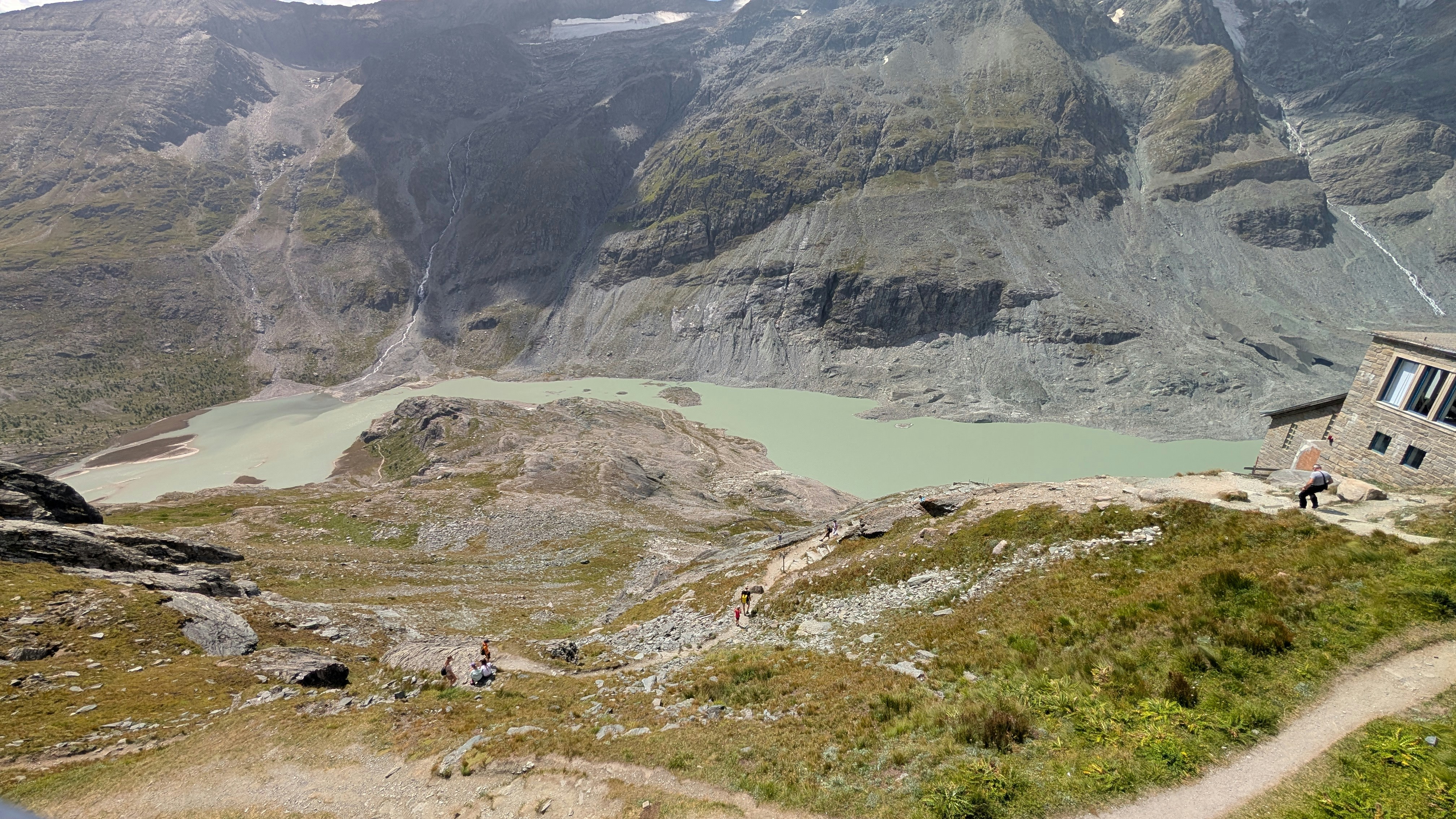 A rugged alpine landscape with a pale turquoise crater lake, rocky slopes, a winding path, and a small building perched near the edge, conveying a remote high-altitude scene.