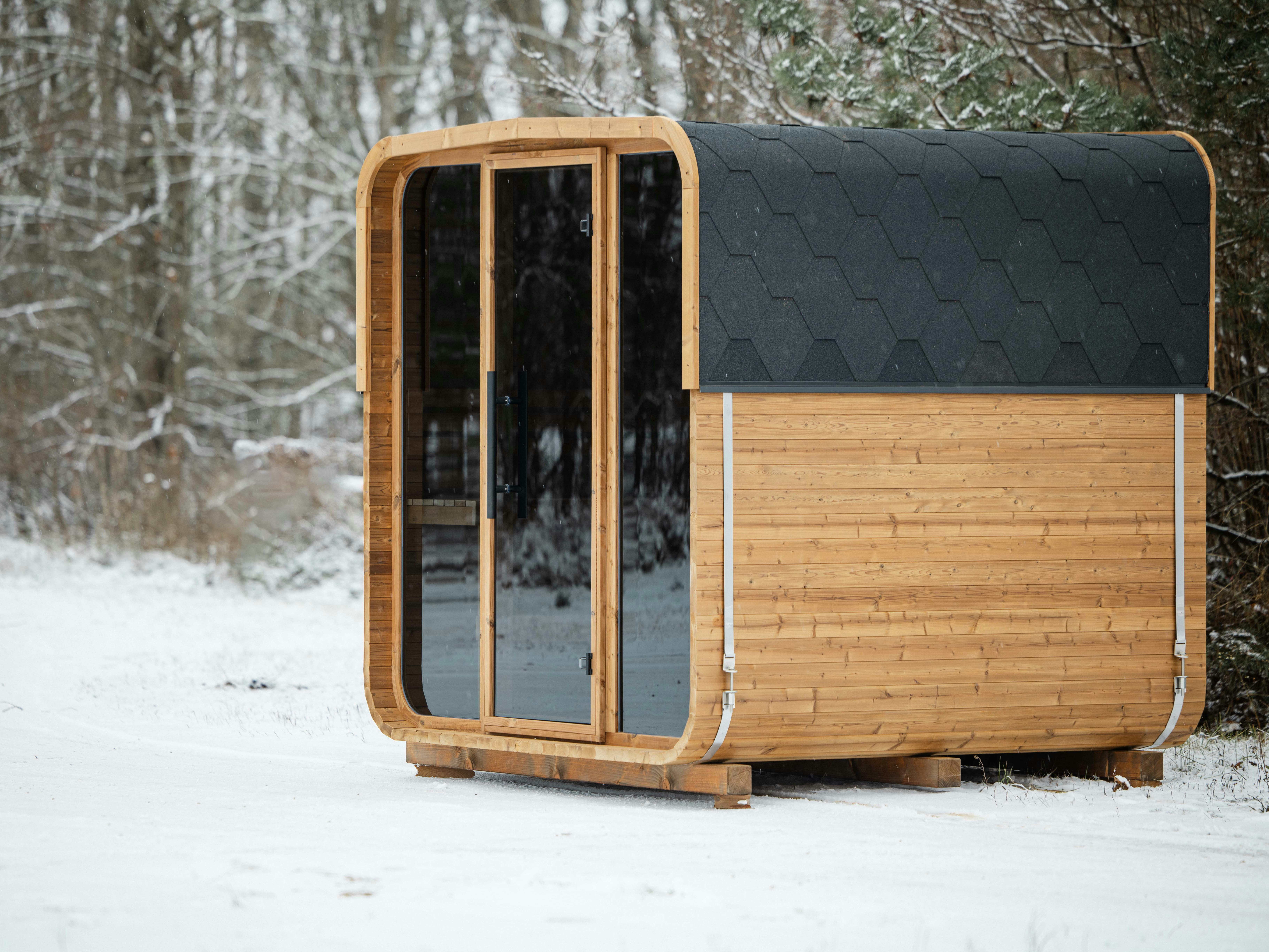 Small wooden cabin with a black hexagonal shingled roof, large glass doors, set in a snowy forest clearing.