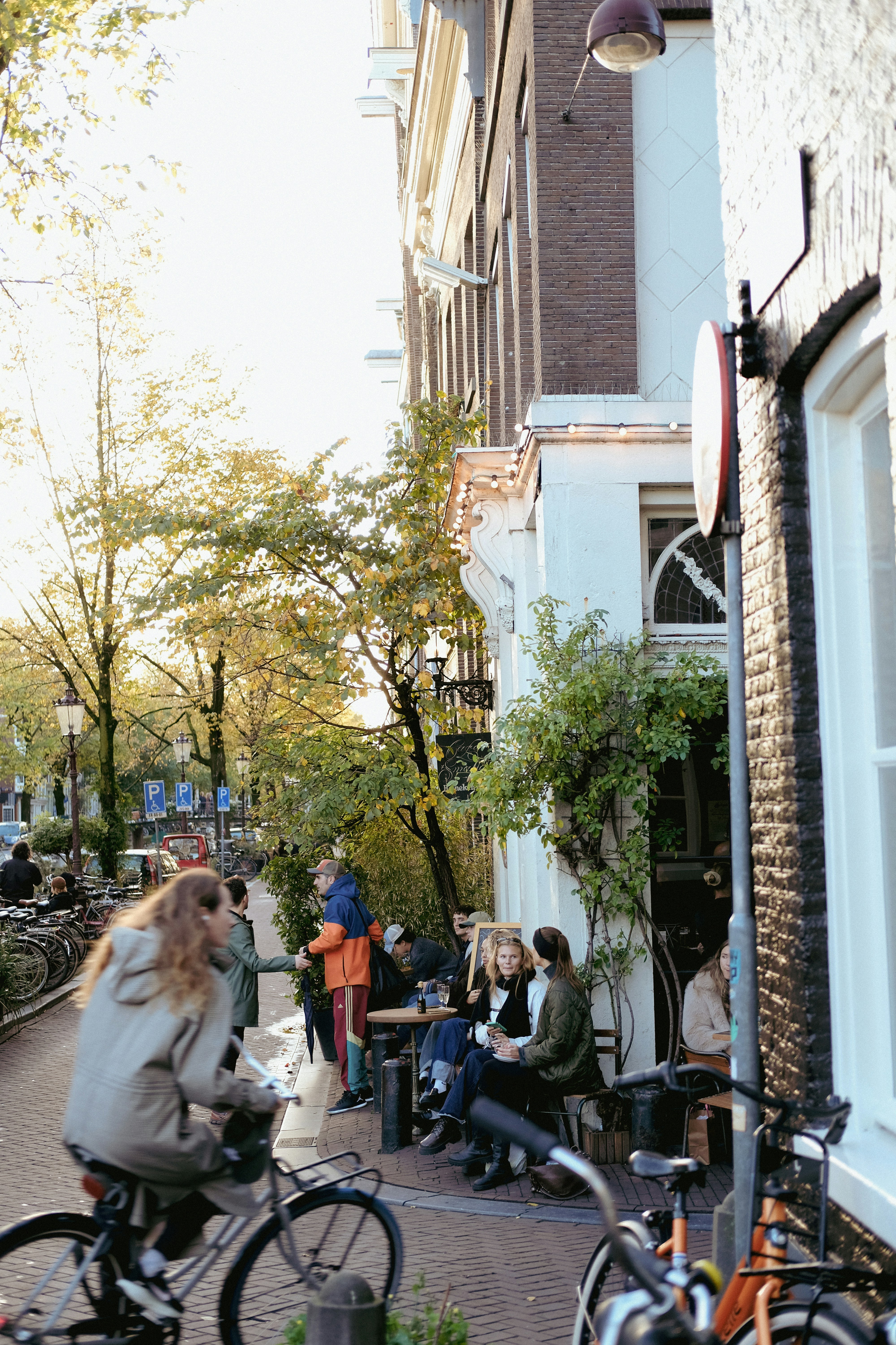 Group of friends enjoying drinks together at an outdoor cafe terrace on autumn day with a cyclist riding past. Candid lifestyle photography showing social gathering, conversation and connection.