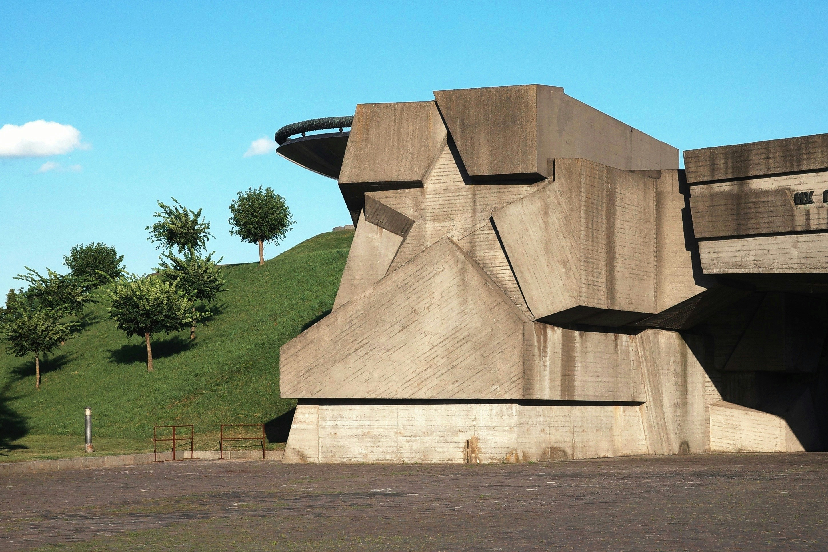 An imposing structure of raw, brutalist concrete, sculpted into angular, geometric forms suggesting a monumental star. This massive, blocky construction rises abruptly from a grassy, sculpted hill planted with small trees, standing in stark contrast against a clear blue sky.