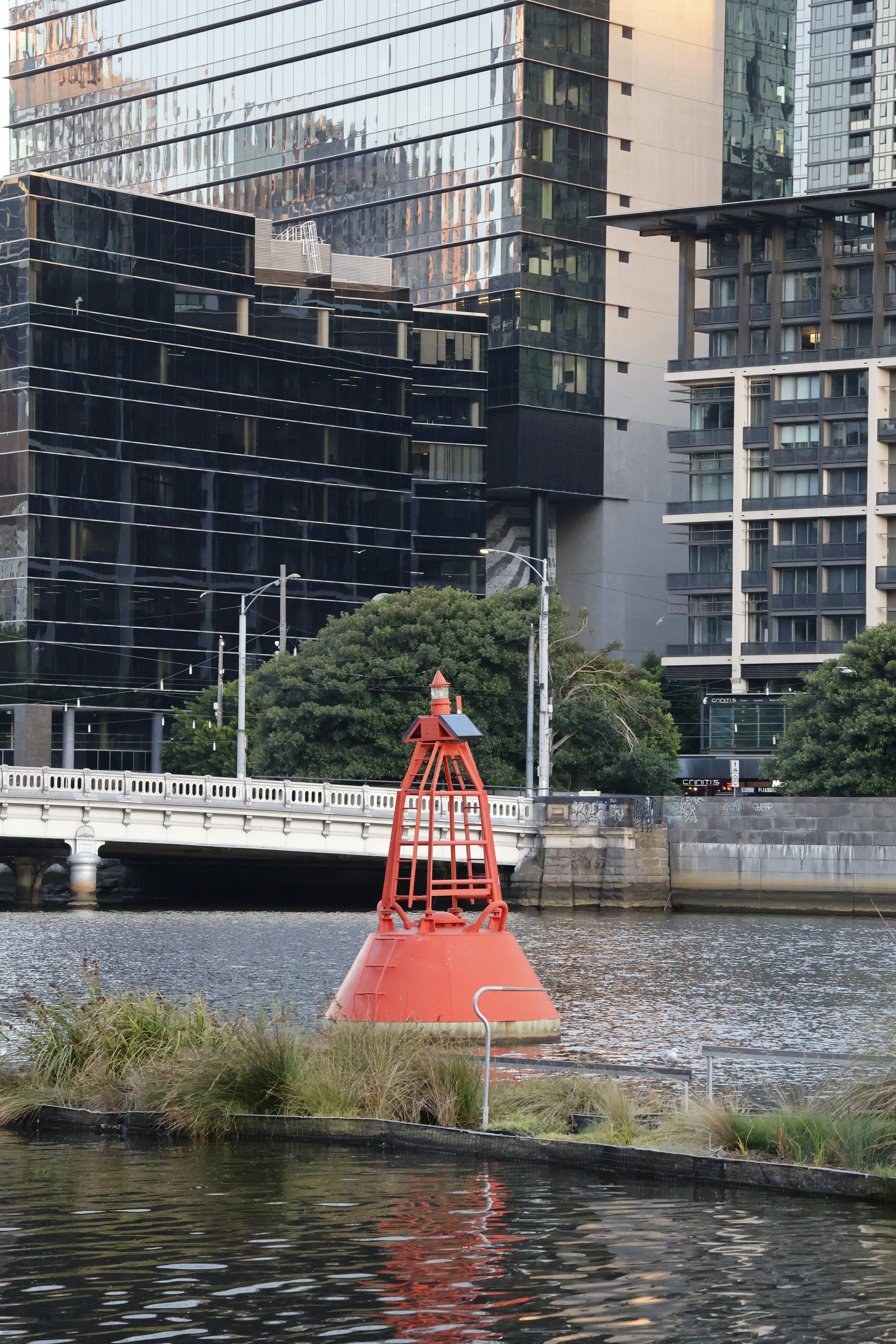 Red beacon against a cityscape: a riverfront sculpture rises from the water