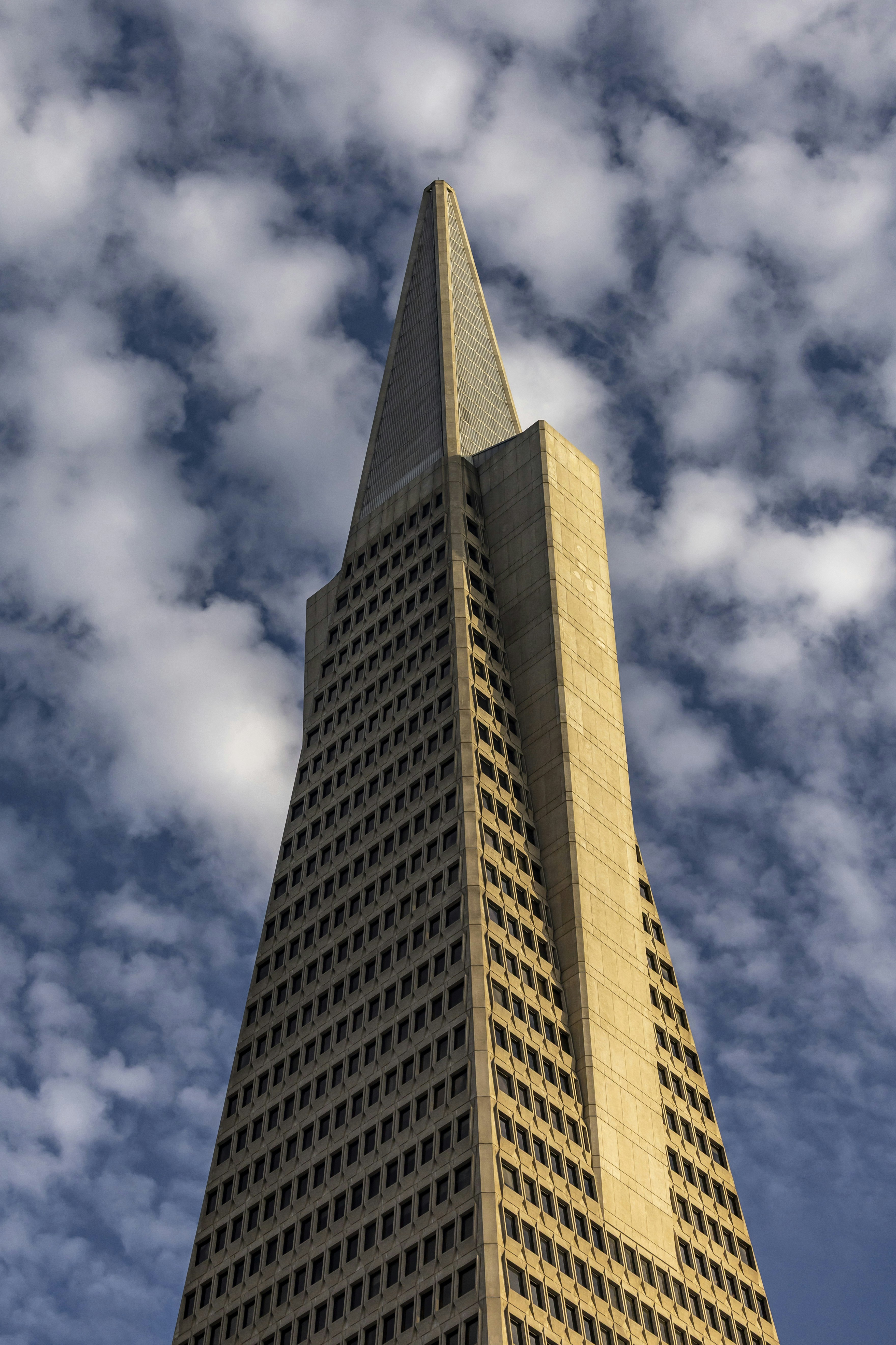 Tall geometric skyscraper piercing a cloudy sky with a warm, sunlit façade, set against a textured blue backdrop. A solitary architectural landmark dominates the frame as clouds drift around.