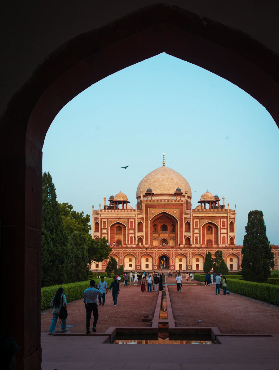People walk toward a grand Mughal monument framed by a dark arch, with a clear blue sky and a reflecting pool at the foreground.