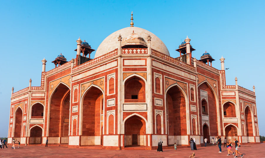Red sandstone and white marble Mughal arches form a grand façade with a central dome in a public square; people provide scale and activity around the architectural monument.