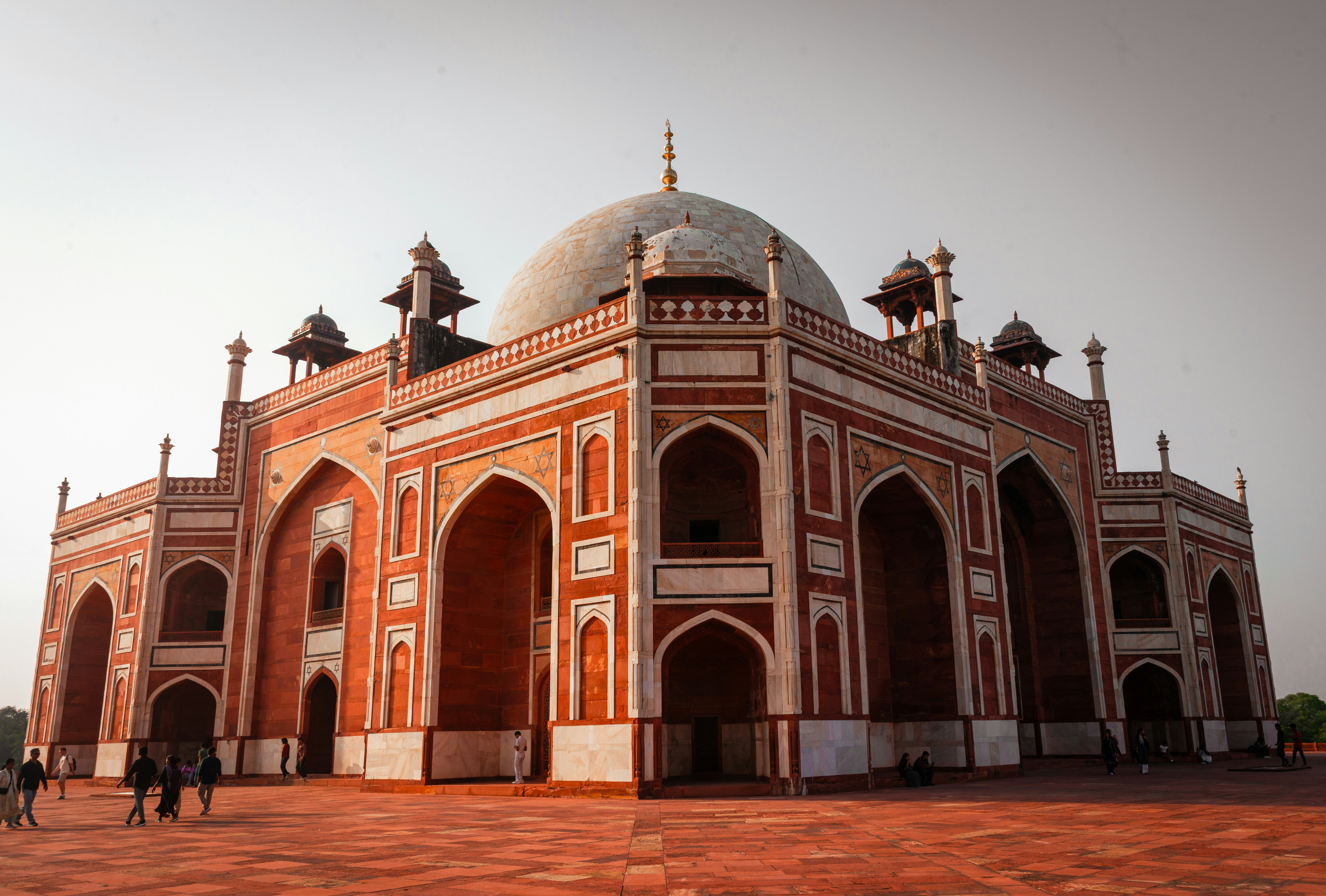 Humayun's Tomb, built by his widow Hamida Banu Begam starting in 1569, is the first distinct example of proper Mughal architecture, influenced by Persian style. Persian architect Mirak Mirza Ghiyath was employed for the project, which cost 15 lakh rupees. It stands centrally within a charbagh (four-part garden) featuring causeways and water channels. The square, double-storeyed red sandstone structure has chamfered corners and is topped by a white marble double dome and pillared kiosks, synthesizing Persian and Indian architectural traditions.