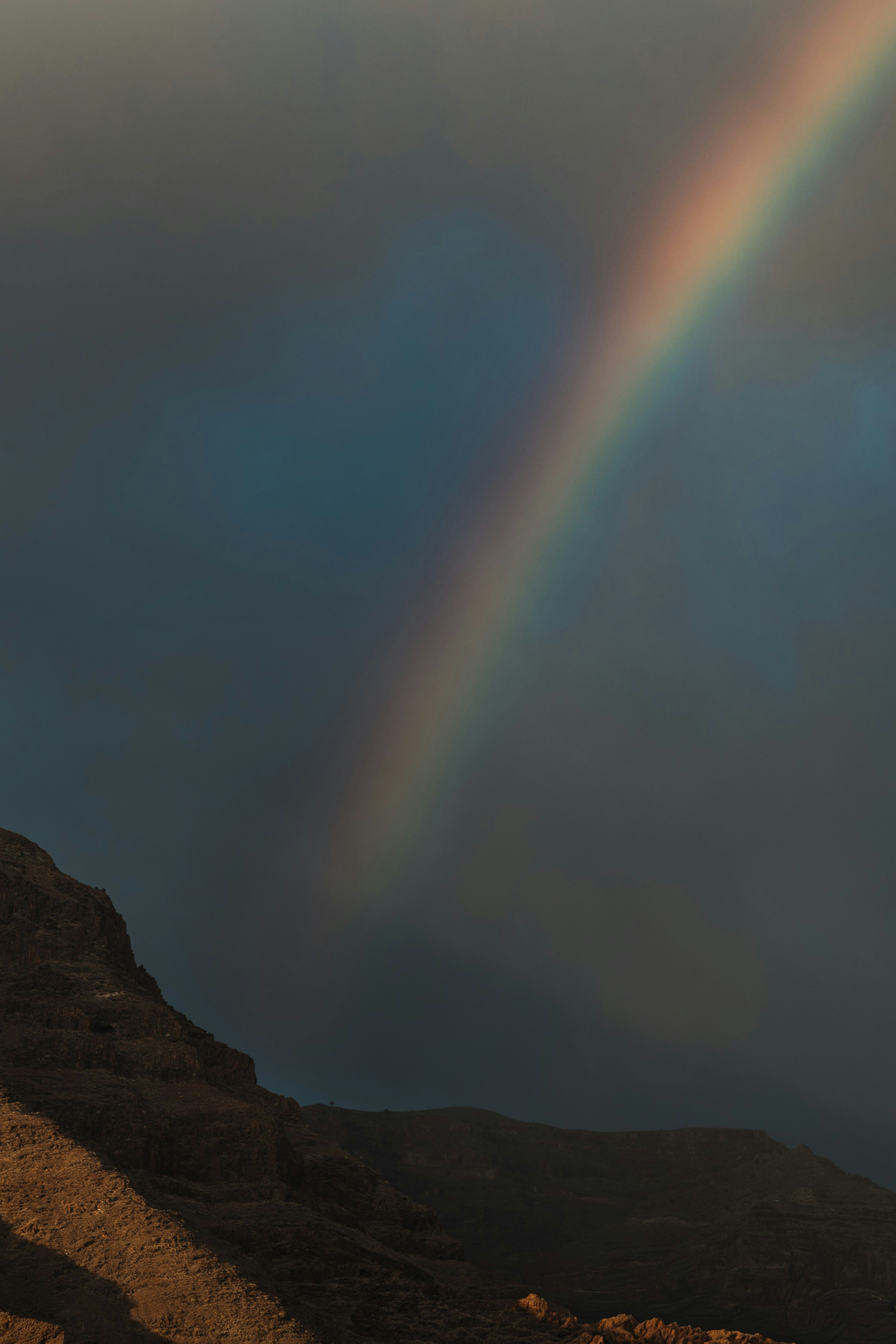 Rainbow emerging over shadowed mountain landscape under dark sky Description: A soft, diagonal rainbow appears against a dark, stormy sky above a rugged mountain range. Warm light on parts of the rocky terrain adds contrast and depth, creating a dramatic and atmospheric scene. Suitable for themes of nature, hope, weather, and scenic wilderness.