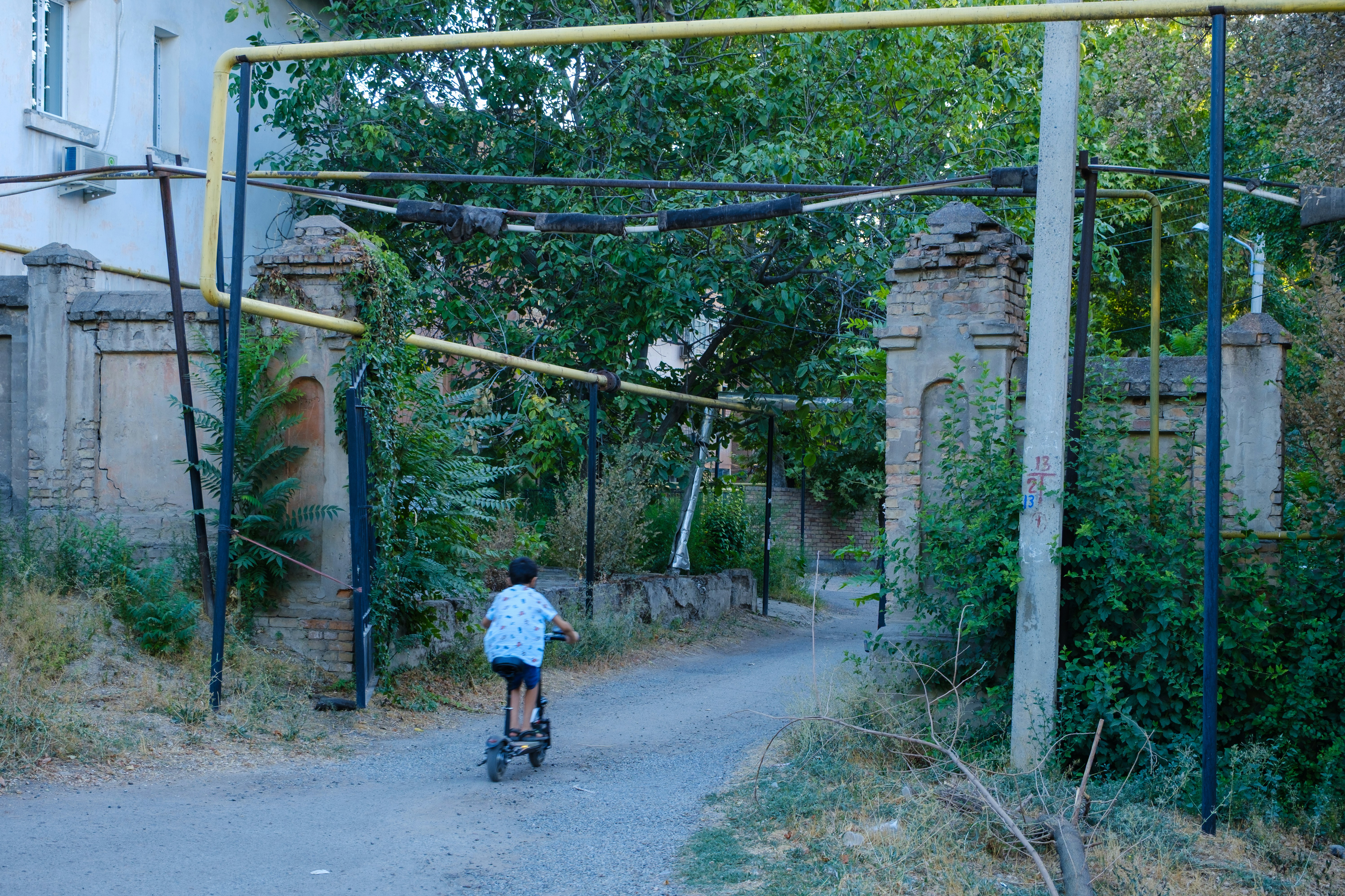Child riding a scooter down a gravel path framed by rustic stone gates and overhanging pipes; a quiet, rural alley with greenery and weathered infrastructure.