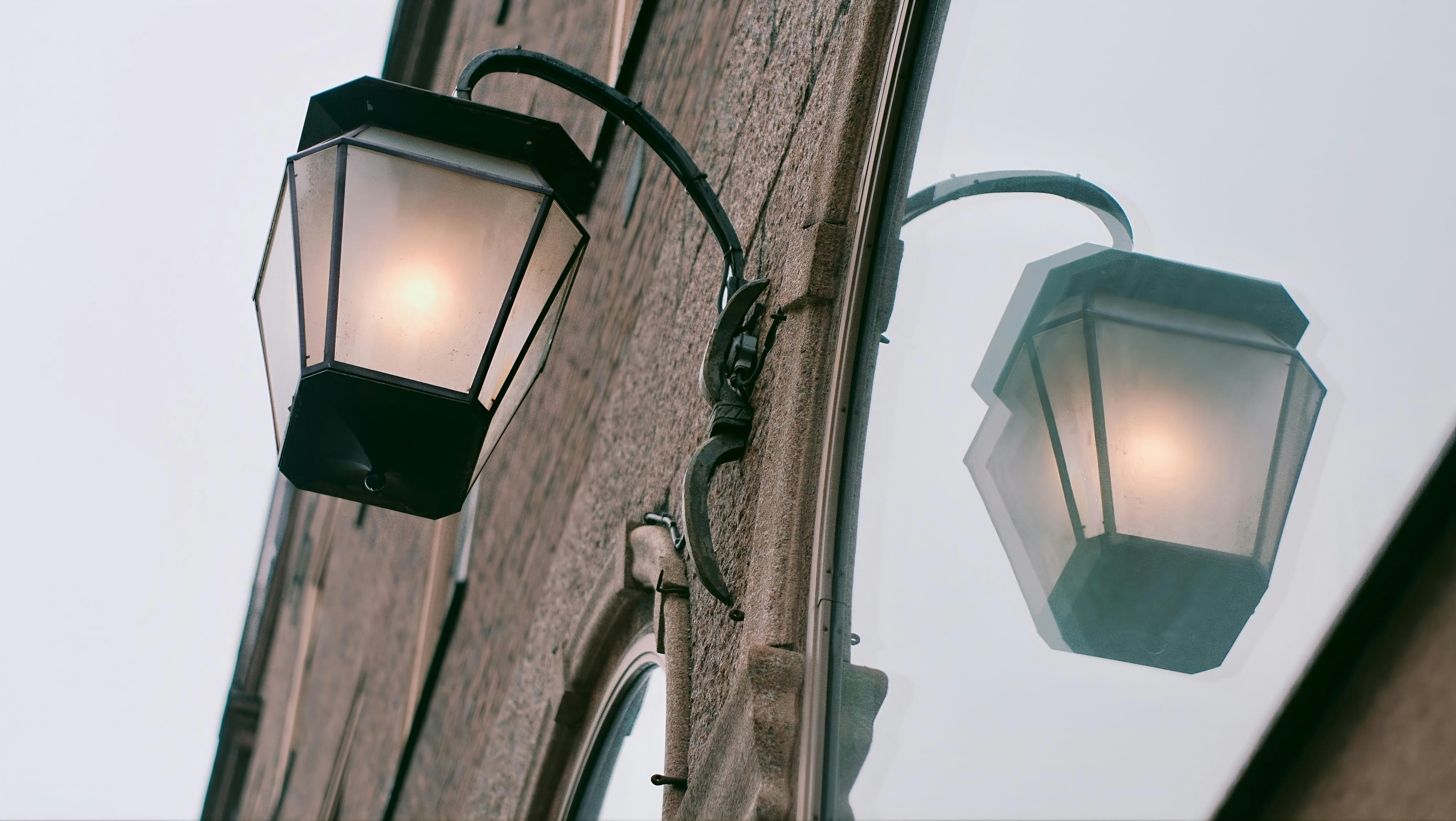 An atmospheric photograph capturing two vintage-style street lamps affixed to an old brick or stone building facade. The hexagonal lanterns, suspended on wrought-iron brackets, glow with a warm, diffuse light through their frosted glass panels. The composition features a striking visual effect where the lamp on the right appears ghosted or double-exposed, overlapping the structure's corner, creating an ethereal and slightly mysterious mood against a bright, overcast sky.