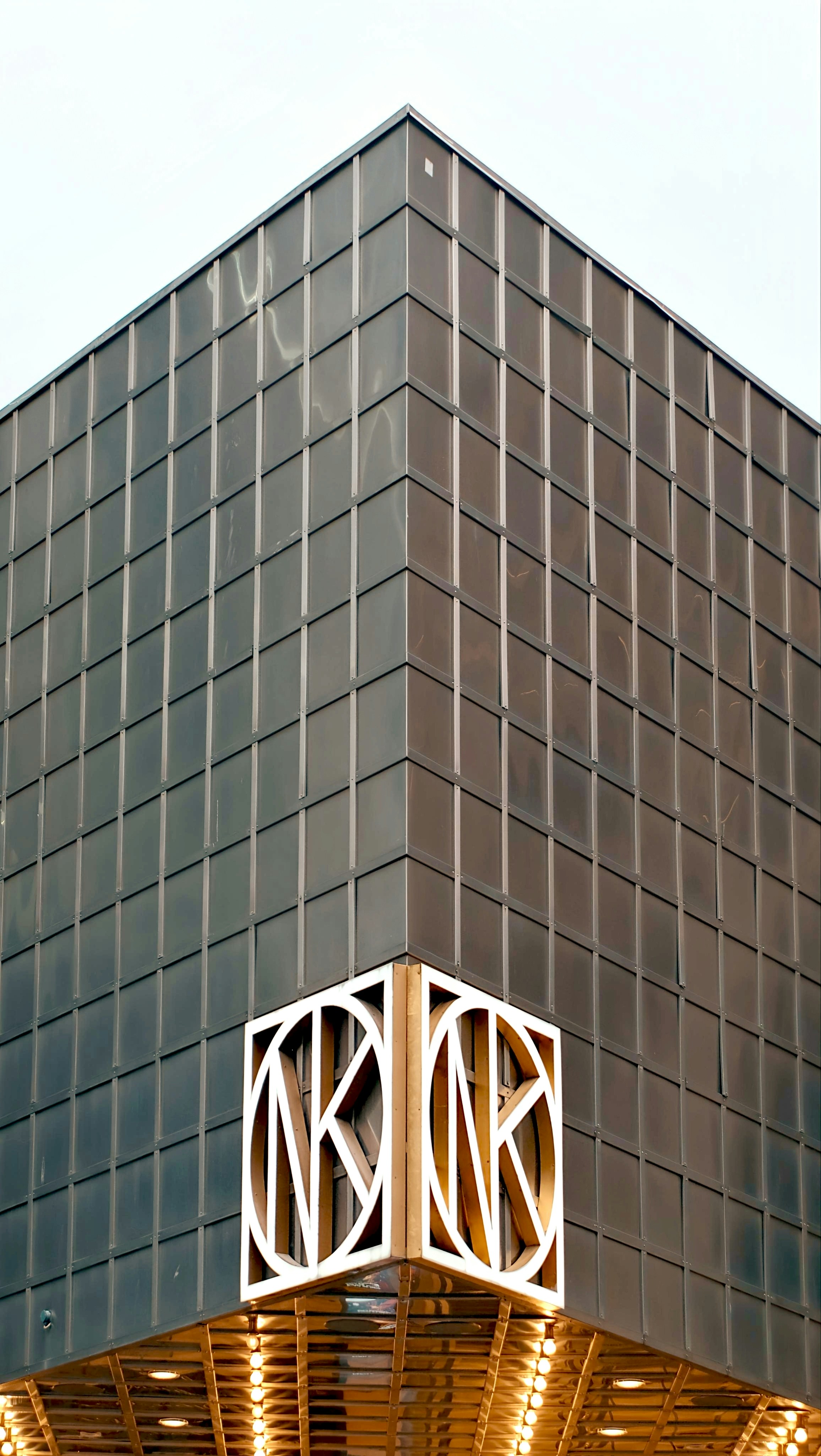 A striking, low-angle photograph focusing on the sharp corner of a modern building. The structure is clad in dark, metallic, grid-patterned panels. Prominently featured at the corner is a large, distinctive square sign with the interlocking white letters "NK" (likely the logo for the department store Nordiska Kompaniet), framed by wooden or gold-toned soffit lighting underneath. The contrast between the dark, gridded surface, the bright white logo, and the warm under-lighting creates a strong, contemporary architectural statement.
