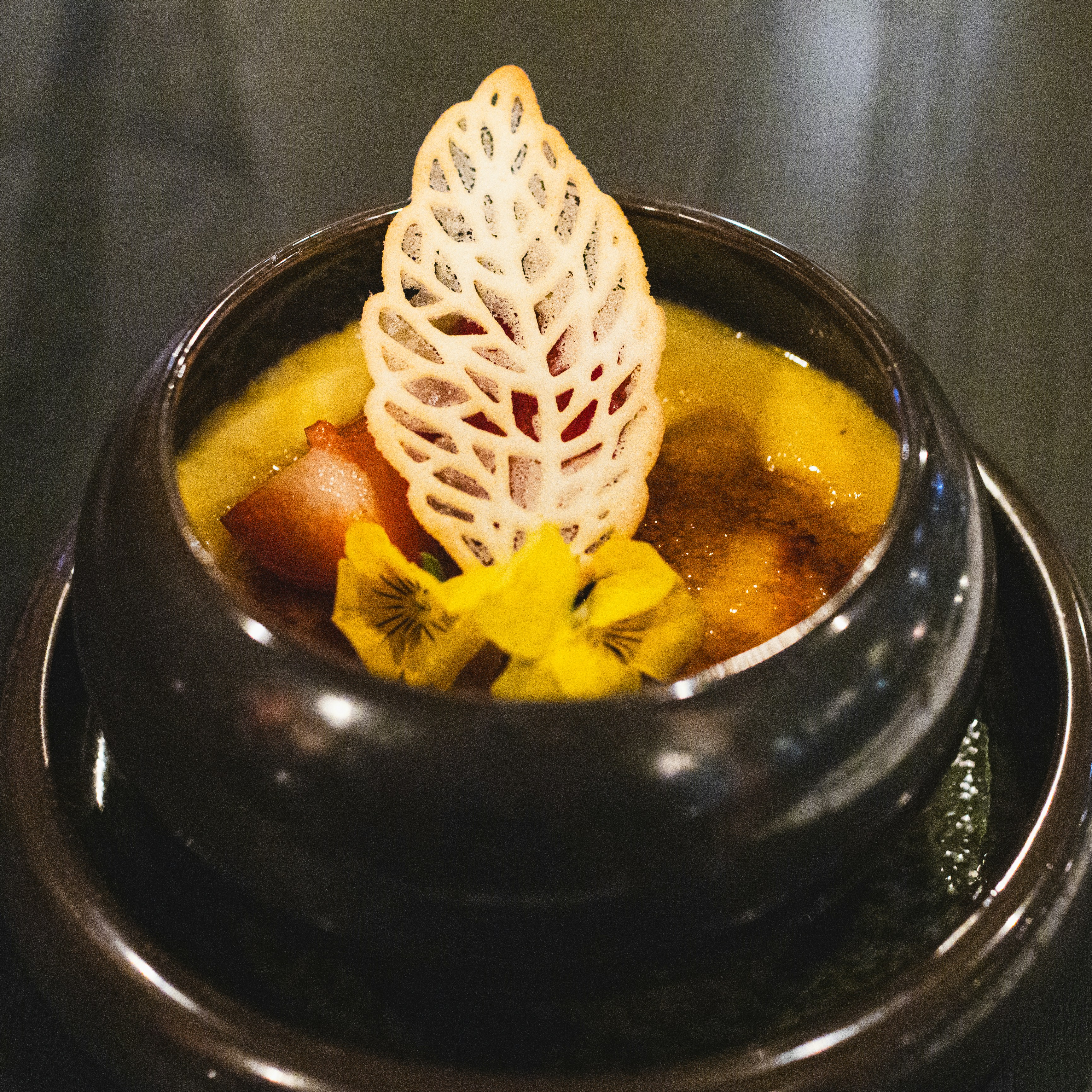 An elegant crème brûlée topped with poached fruit, yellow pansies, and a delicate lattice tuile in a glossy black ceramic bowl at MOMI West-African fusion restaurant in Diemen.