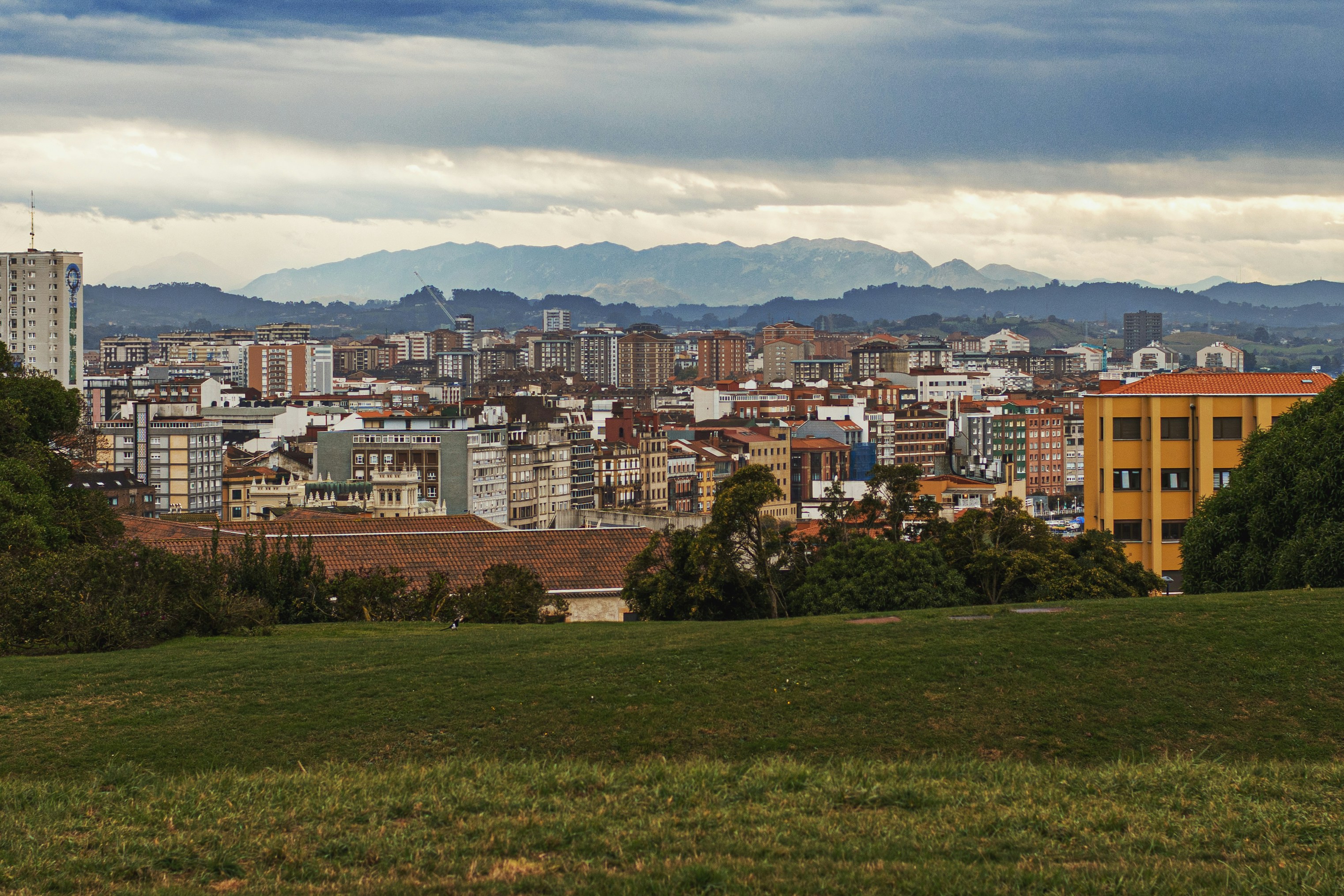 The rooftops of Gijón spread across the foreground as the Cantabrian Mountains rise dramatically in the misty distance—urban Asturias meets wild peaks.