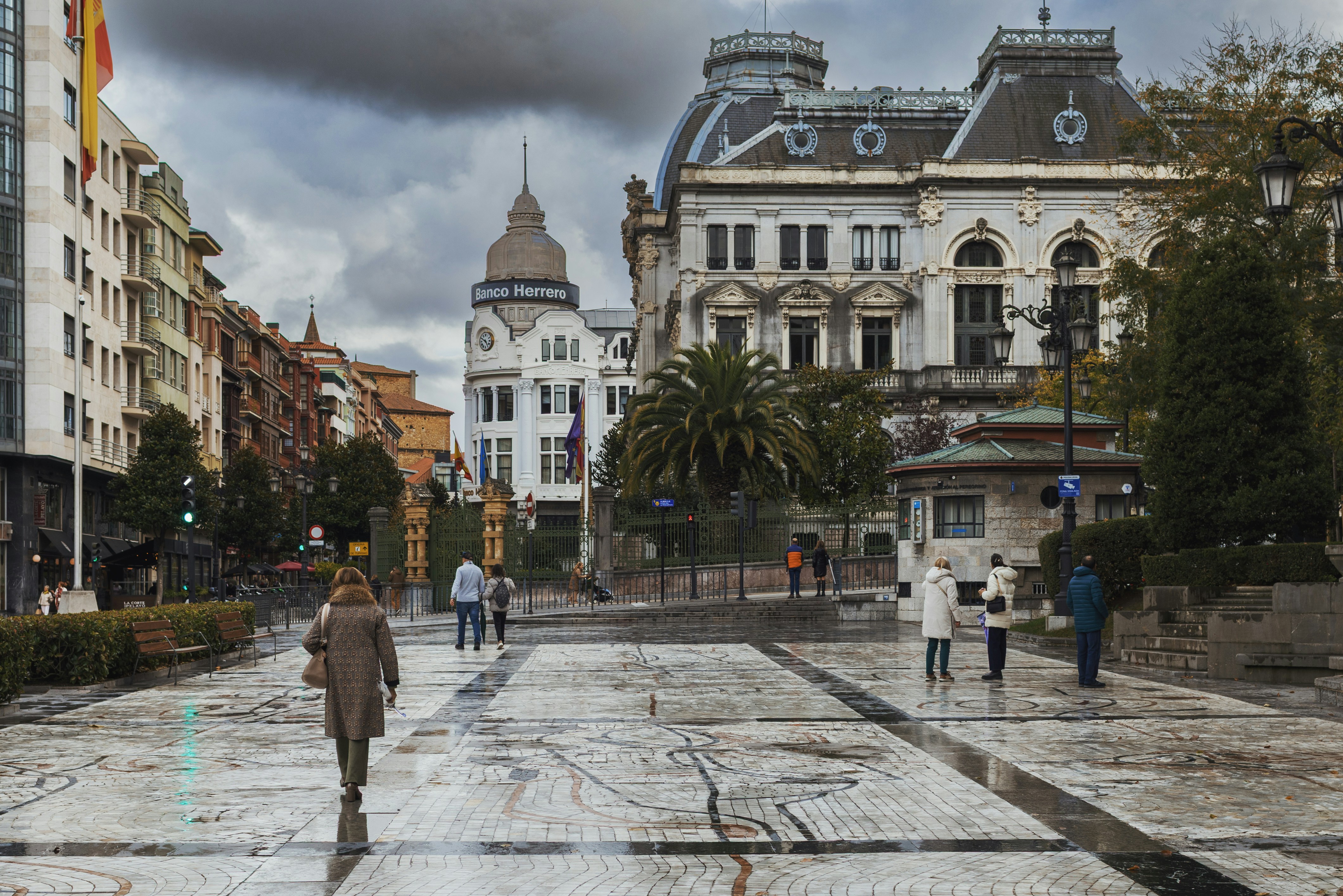 Pedestrians cross the wet marble plaza near the ornate Banco Herrero building in Oviedo, dramatic clouds gathering over the historic Asturian architecture.