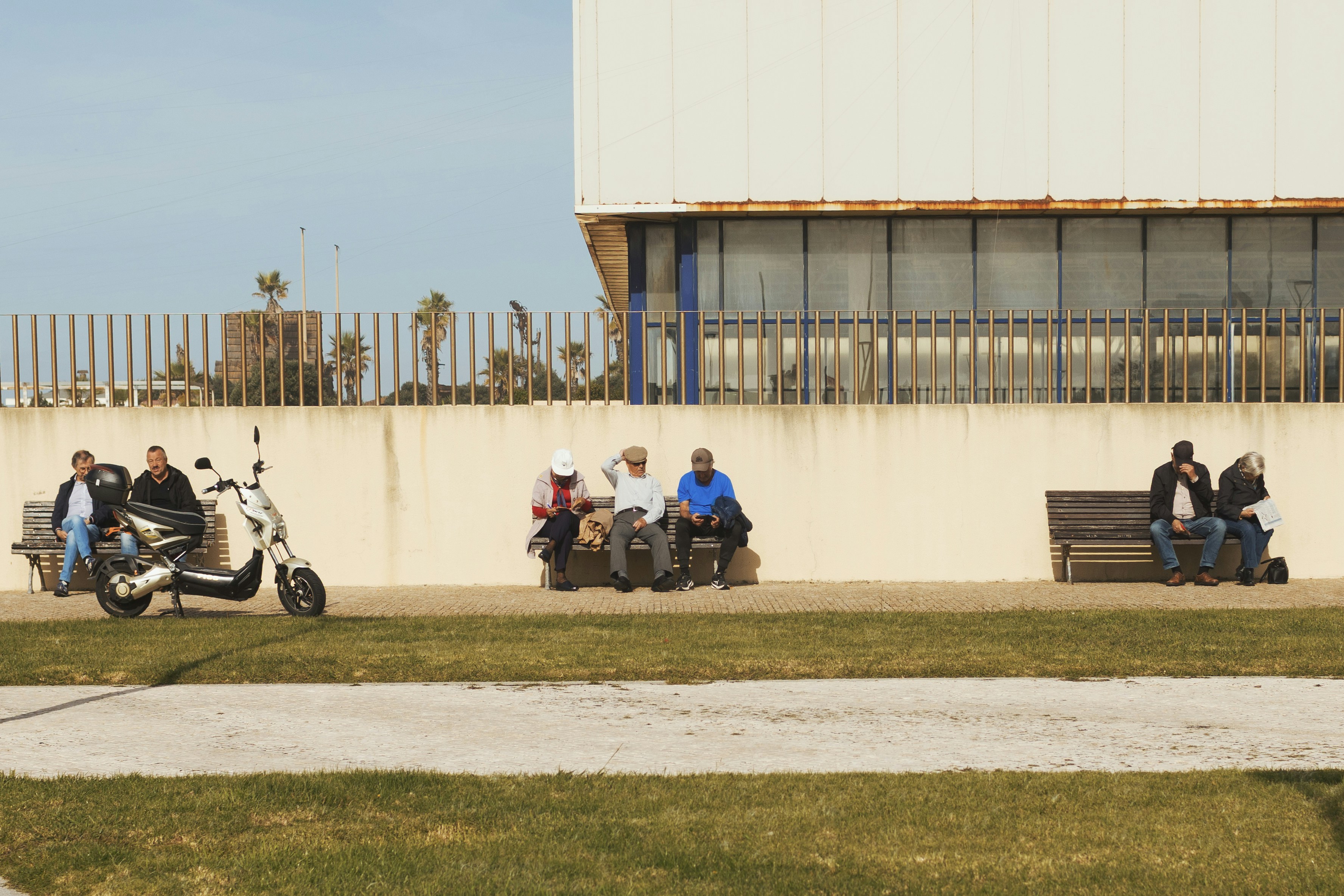 Locals relax on benches beneath a modernist building at Póvoa de Varzim beach, palm trees and clear skies suggesting a mild autumn day on the Portuguese coast.