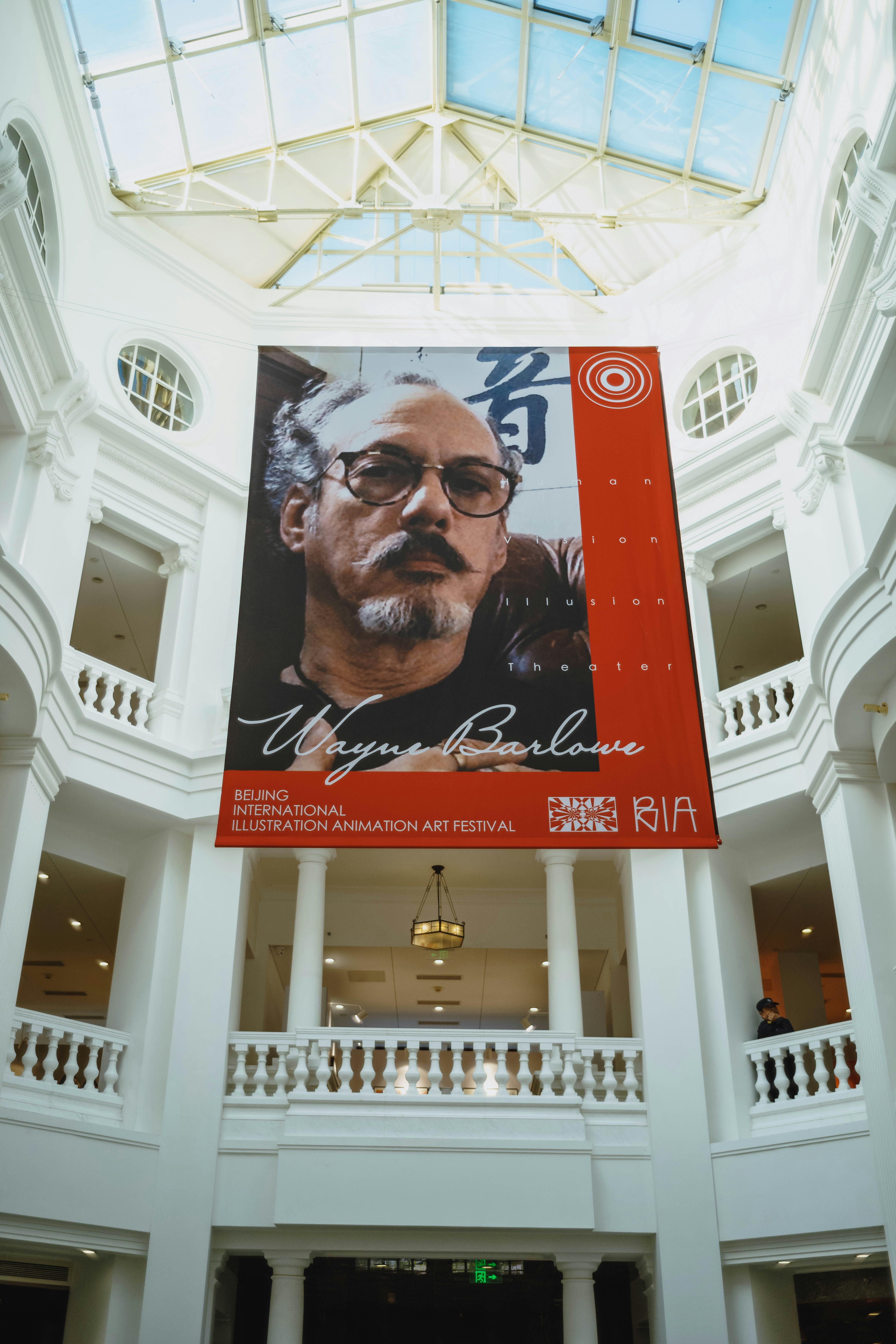 Banner Portrait: A towering red poster of Wayne Barlow dominates a sunlit atrium
