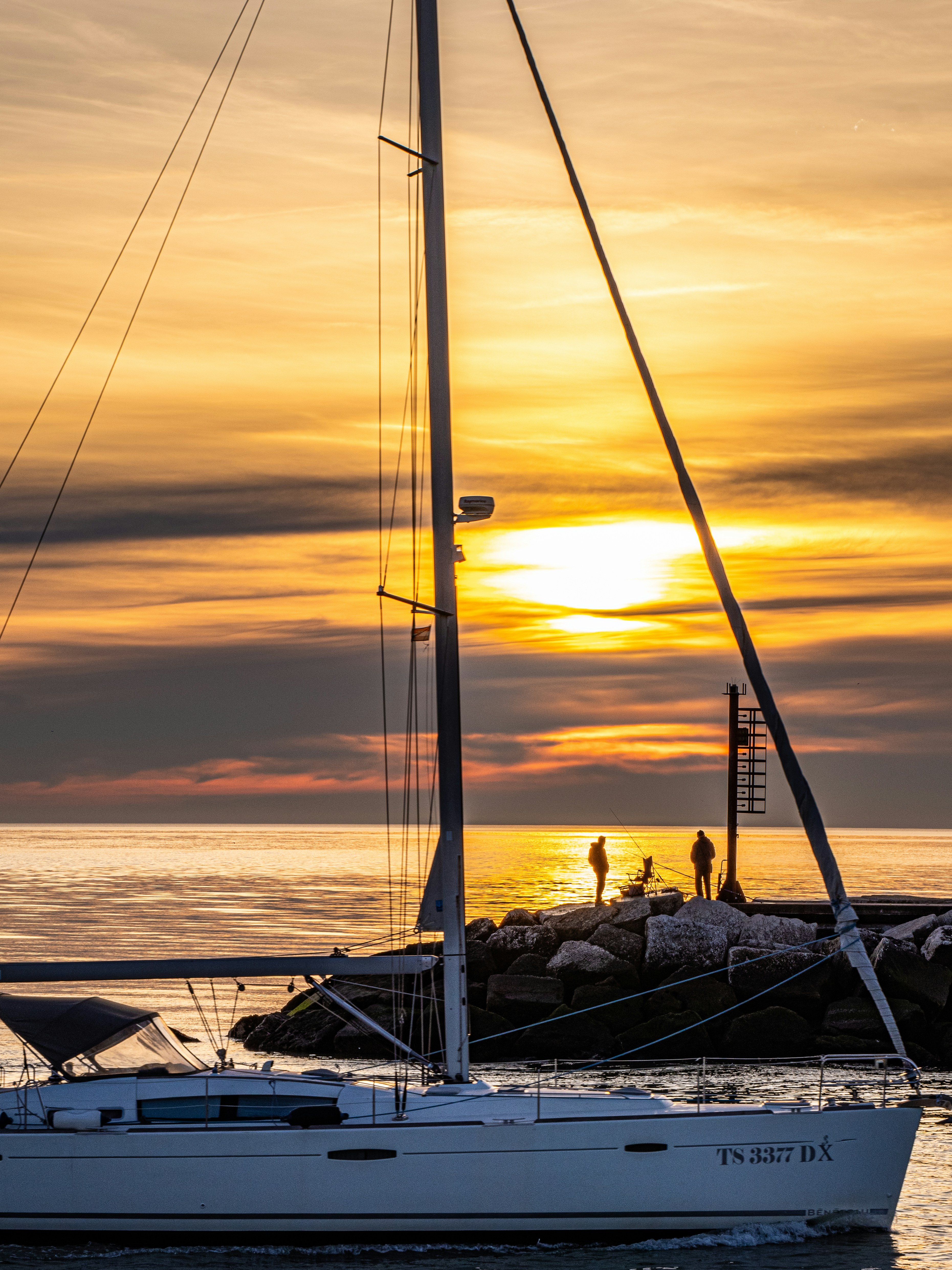 Sailboat at Sunset: Figures on Breakwater Beneath Golden Sky