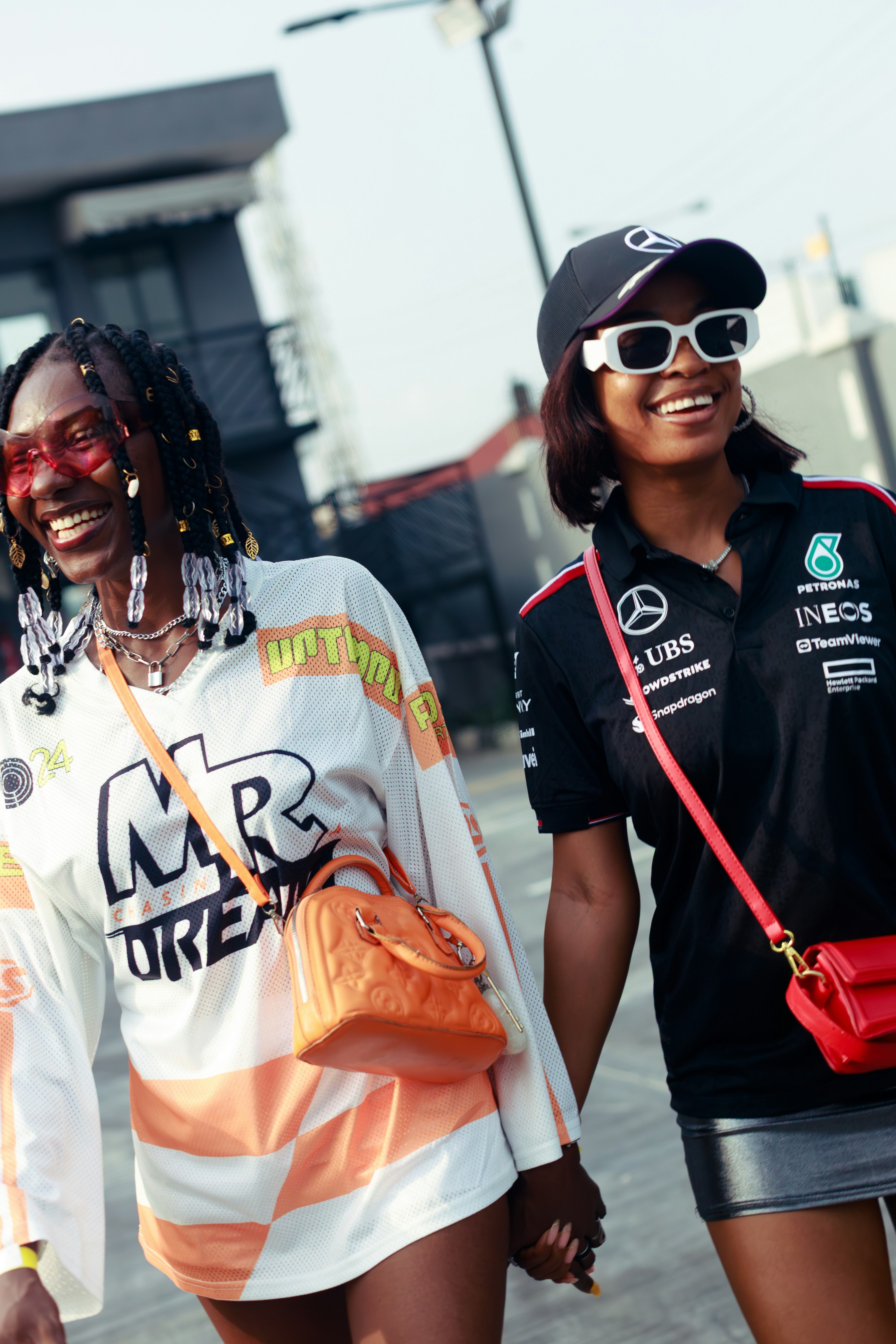 Two friends in vibrant street wear share a bright smile under a sunny urban backdrop