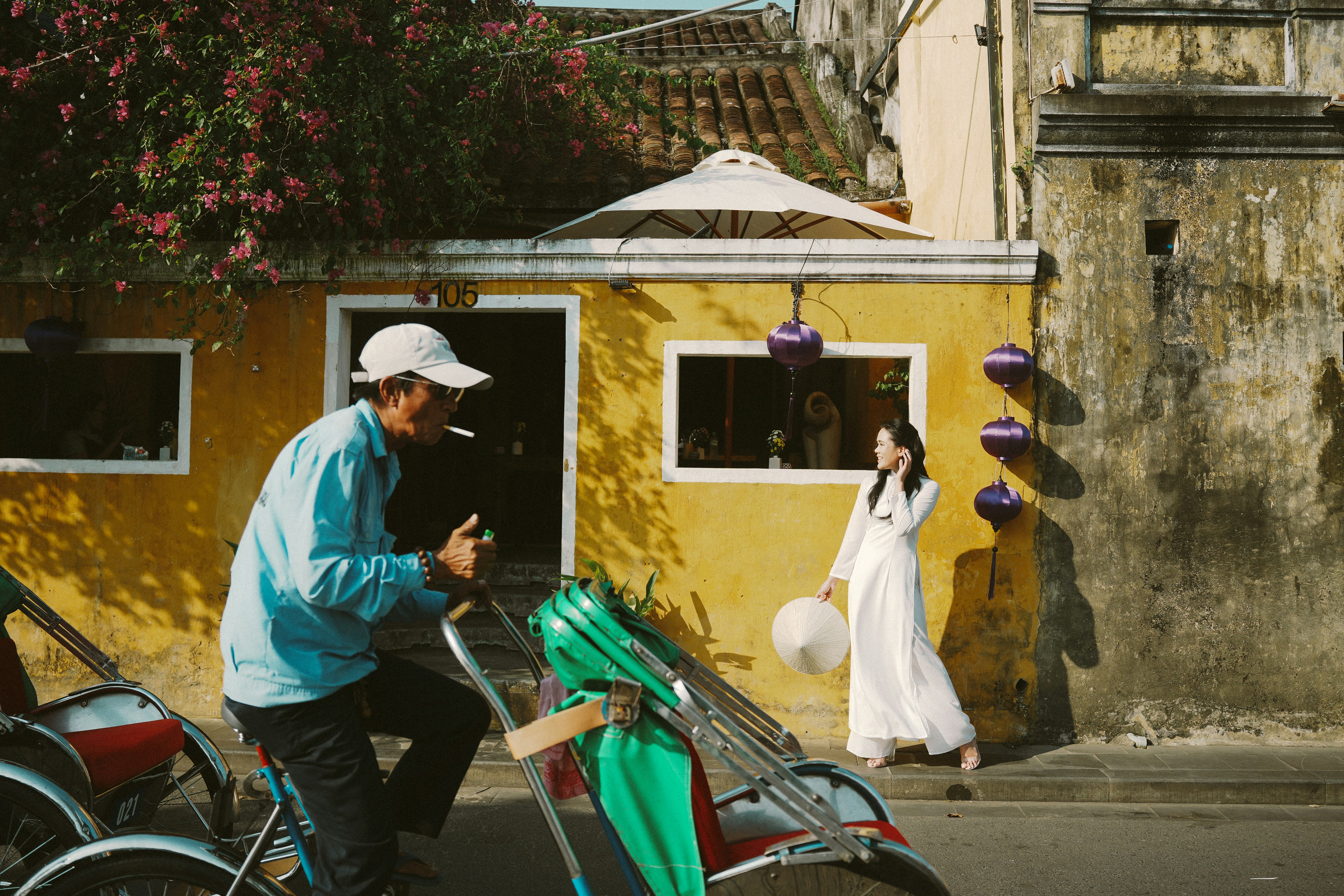 Yellow storefront, passing cyclist, and a distant bride: a candid street moment in tropical light