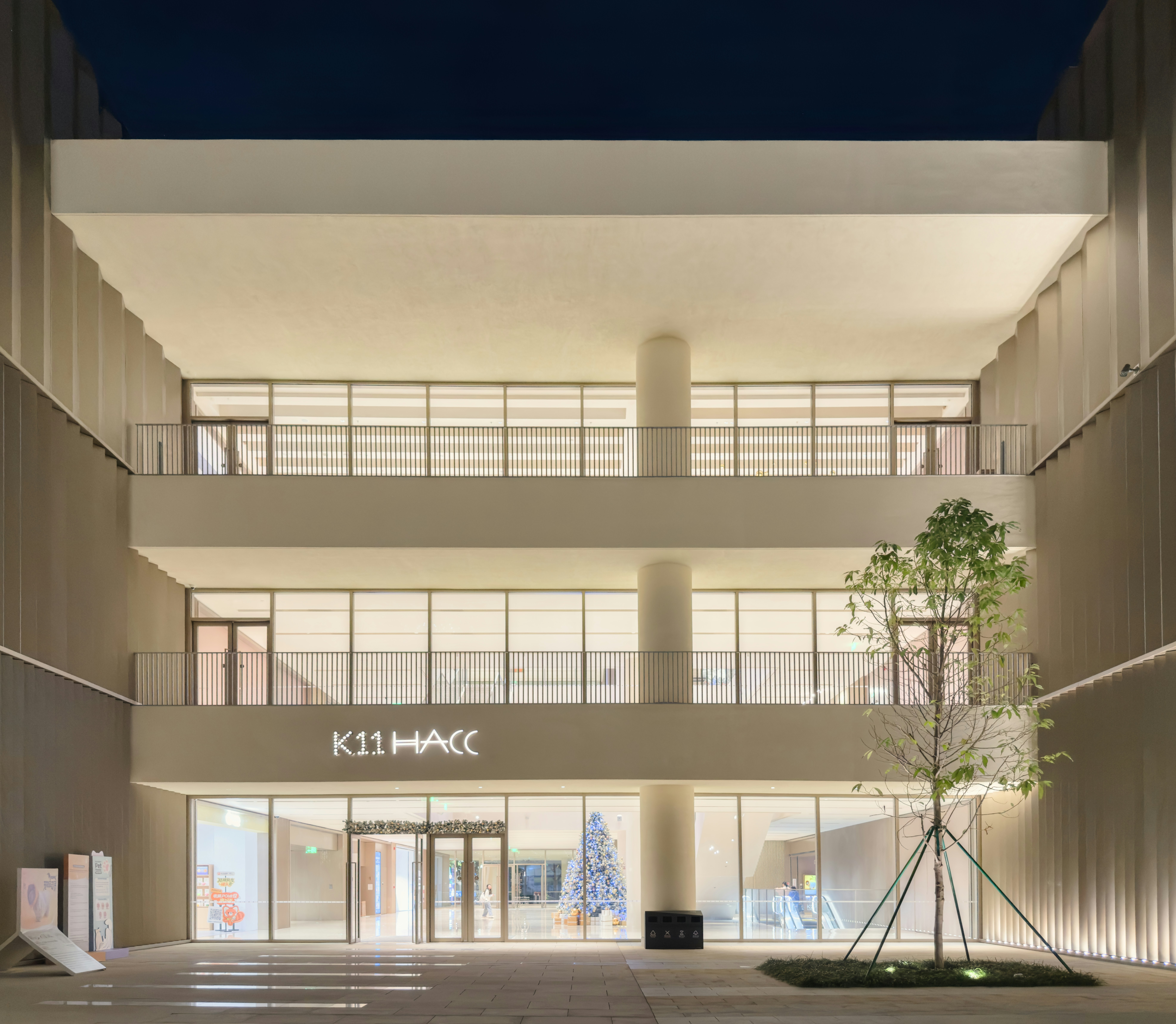 A clean, modern architectural facade captured at night at K11 HACC. Soft warm interior lights illuminate the geometric lines of the balconies and pillars, while a Christmas tree adds a subtle festive touch in the background. The symmetry, open space, and minimal aesthetic highlight contemporary urban design.