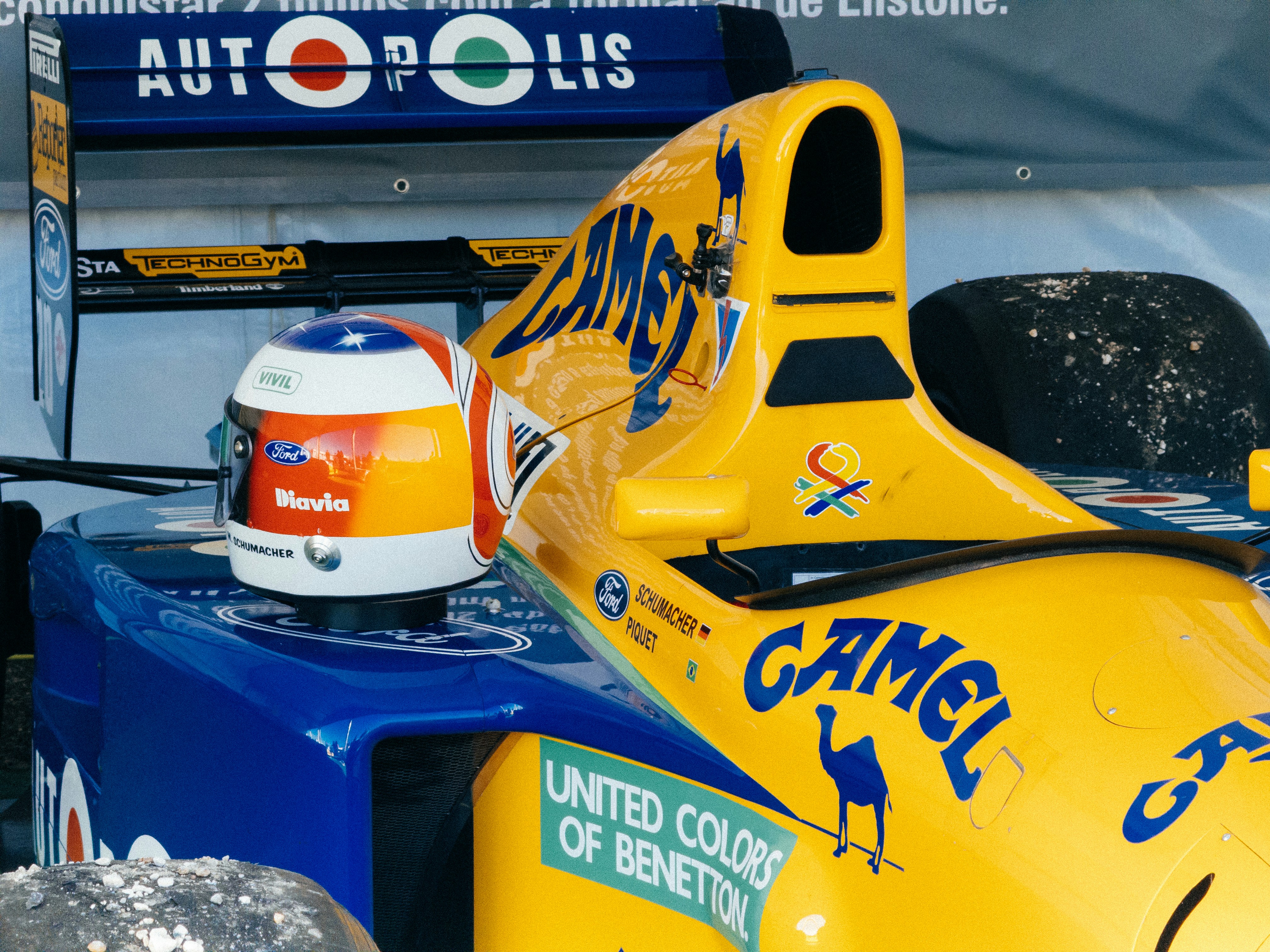 Close-up of a bright yellow Formula 1 car with a blue nose section and a white and orange racing helmet resting on top, parked in a pit area with branding visible on the bodywork.