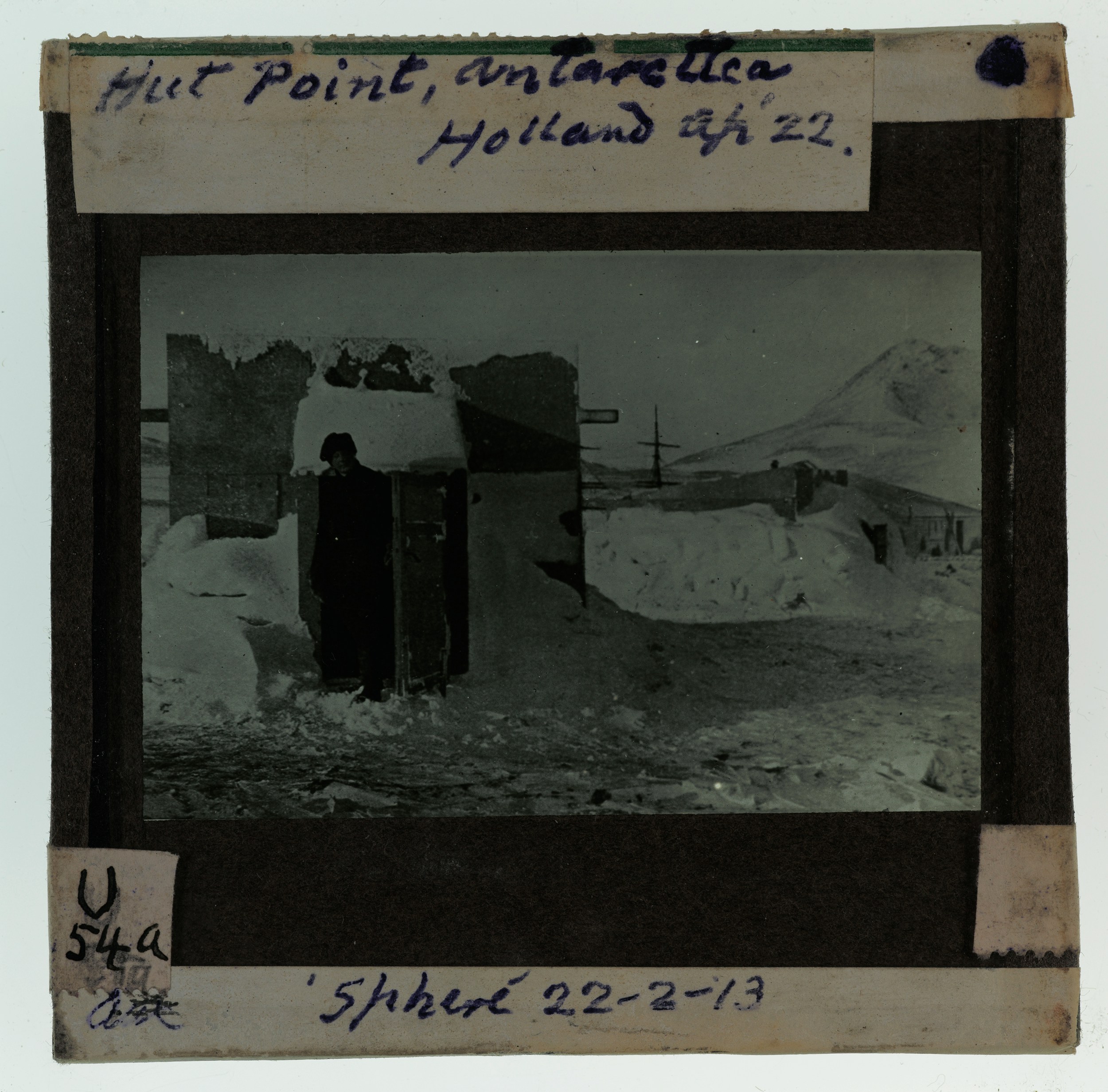 A lantern slide produced by Clement Lindley Wragge (b.1852, d.1922), dated 22 February 1913. The slide shows the image of a heavily cloaked person emerging from a small building covered in snow. The ground is also covered in snow. In the background is a ship's mast and other larger structures. Beyond that is a large mountain. Shared by Auckland War Memorial Museum, Tāmaki Paenga Hira, as part of the Clement Lindley Wragge collection.