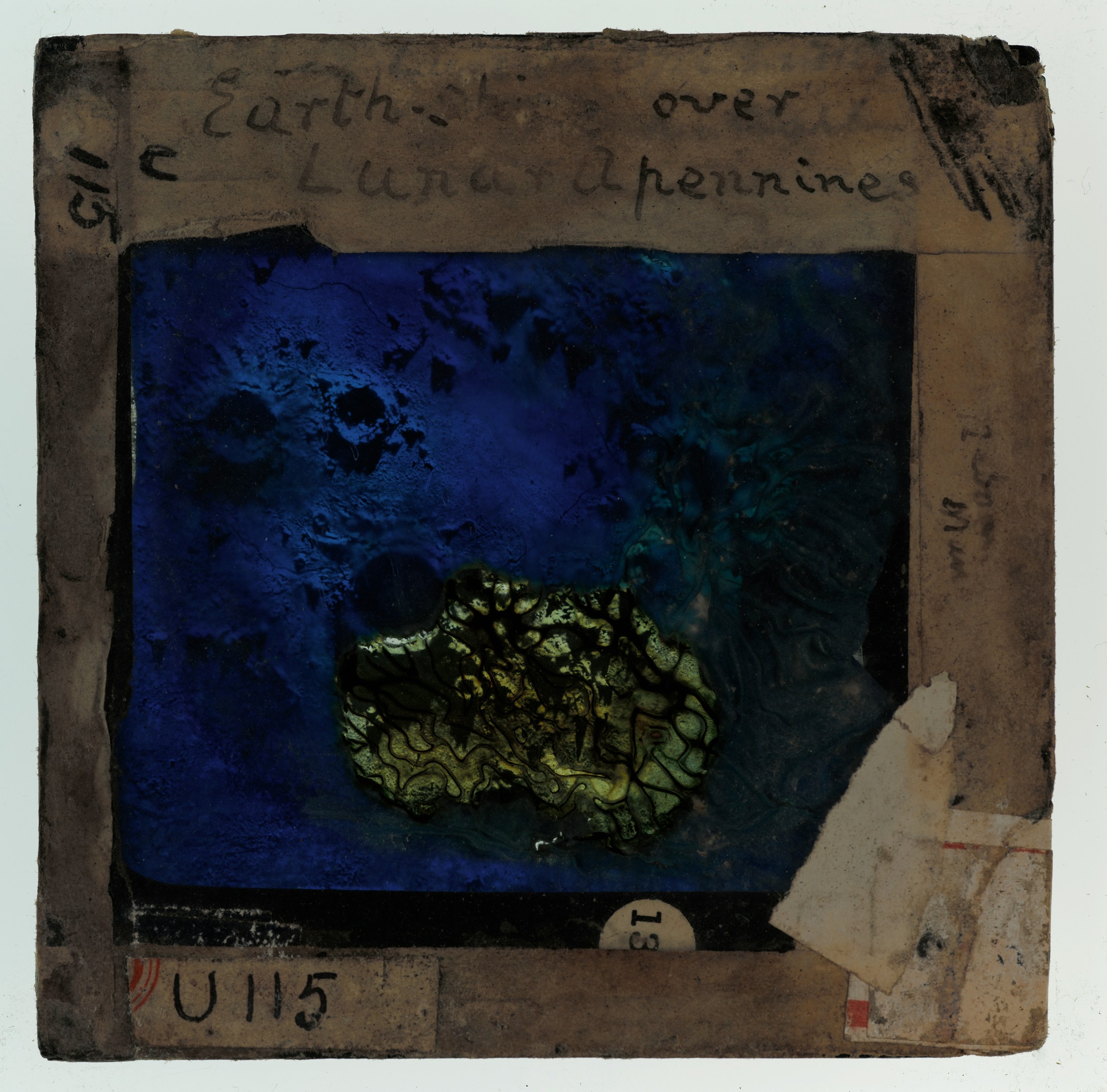 A textured, square frame houses a deep blue central area with a glowing, cracked-yellow island-like texture, suggesting a celestial or geological motif; the piece appears to be an art print or mixed-media work with visible wear and handwritten notes along the border.