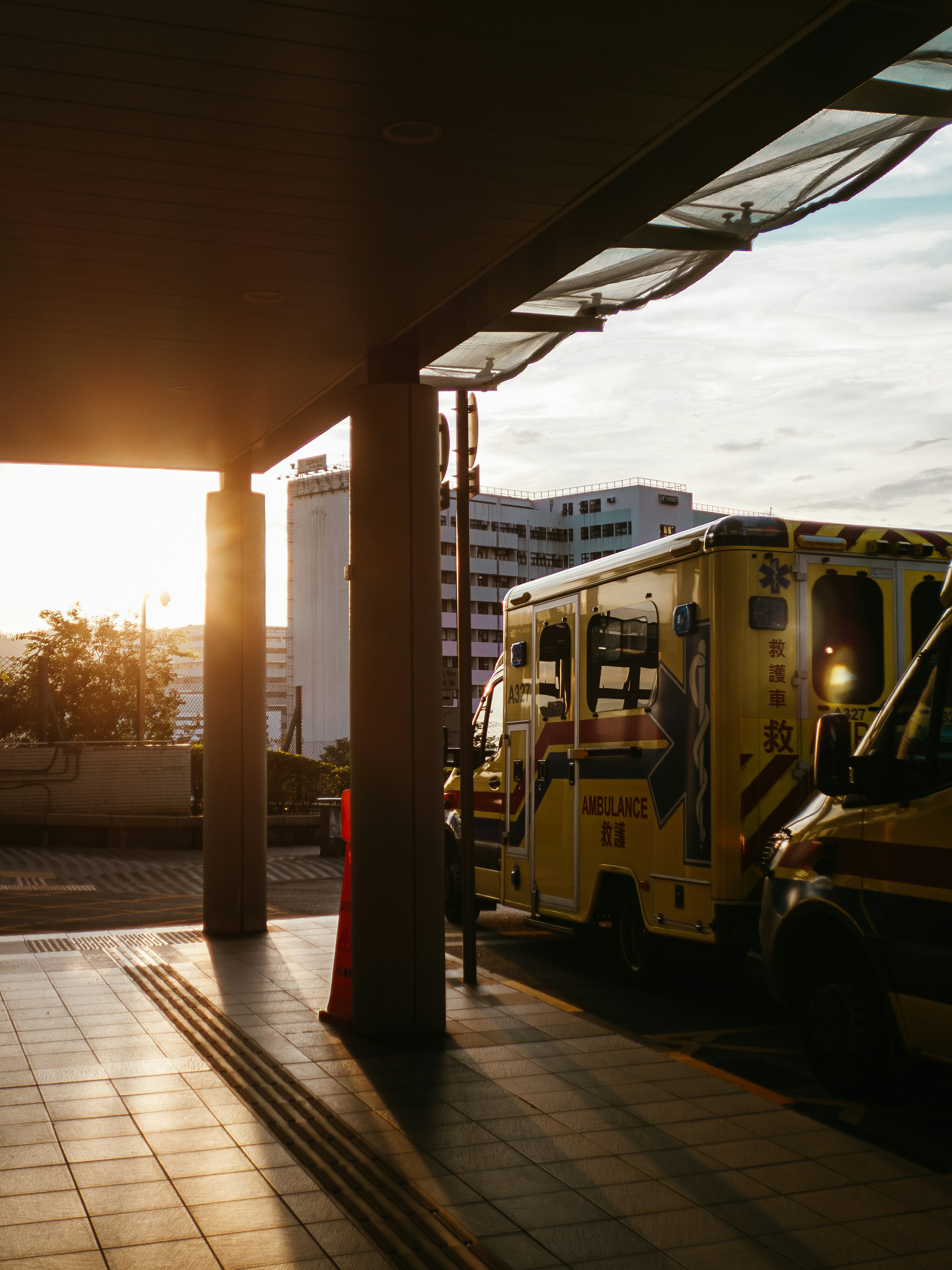 The golden hour casts a serene glow over the entrance of Kwai Chung Hospital in Hong Kong, illuminating a parked yellow ambulance in warm, amber light. Long, dramatic shadows stretch across the tiled walkway as the sun dips low, silhouetting the building's sturdy pillars.