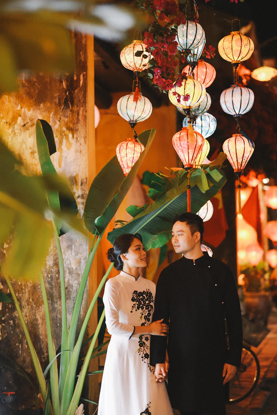 Couple holding hands beneath hanging lanterns and tropical foliage in a warm night market scene.