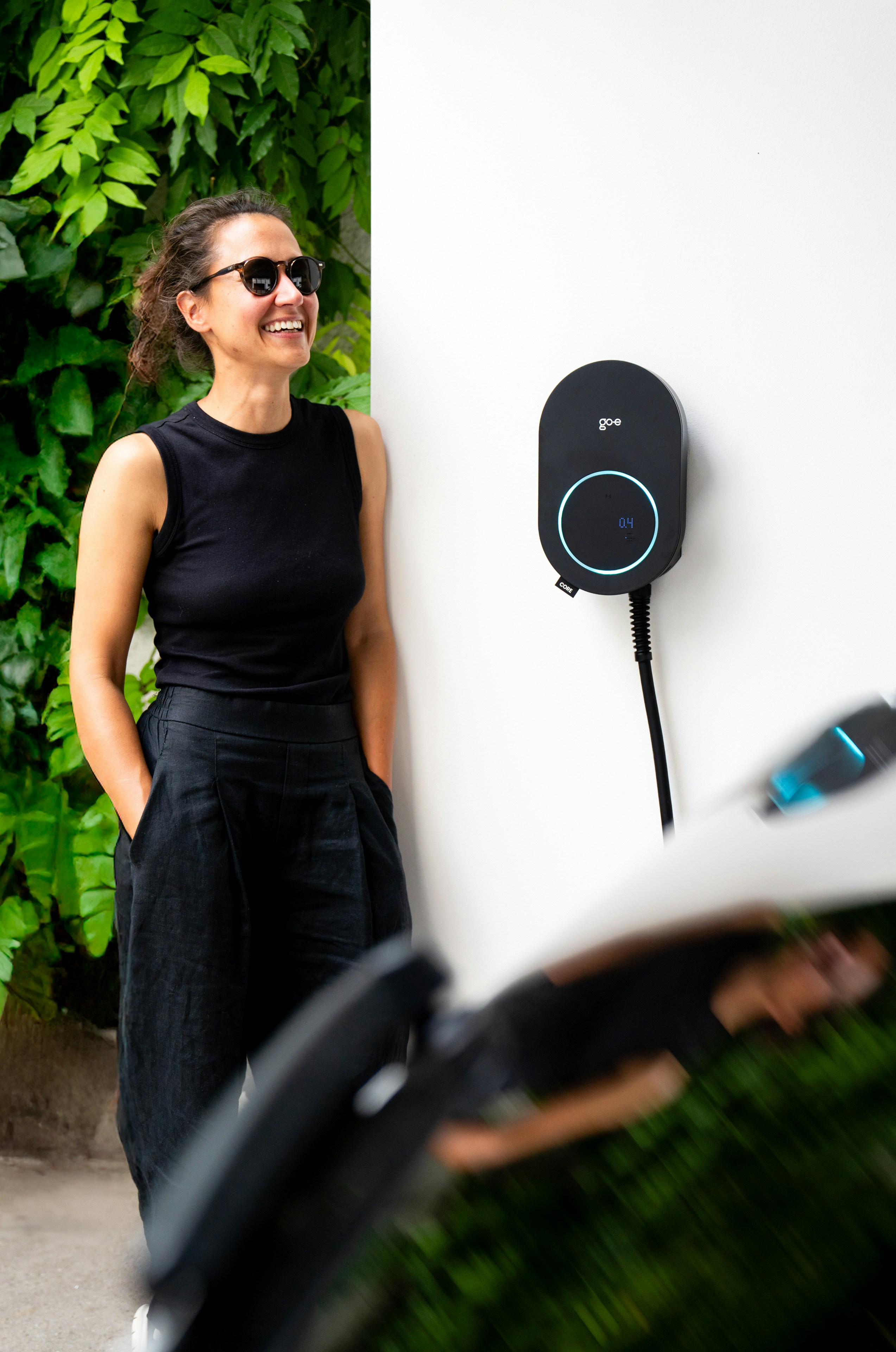 A woman stands by a wall-mounted go-e Charger CORE under green vines